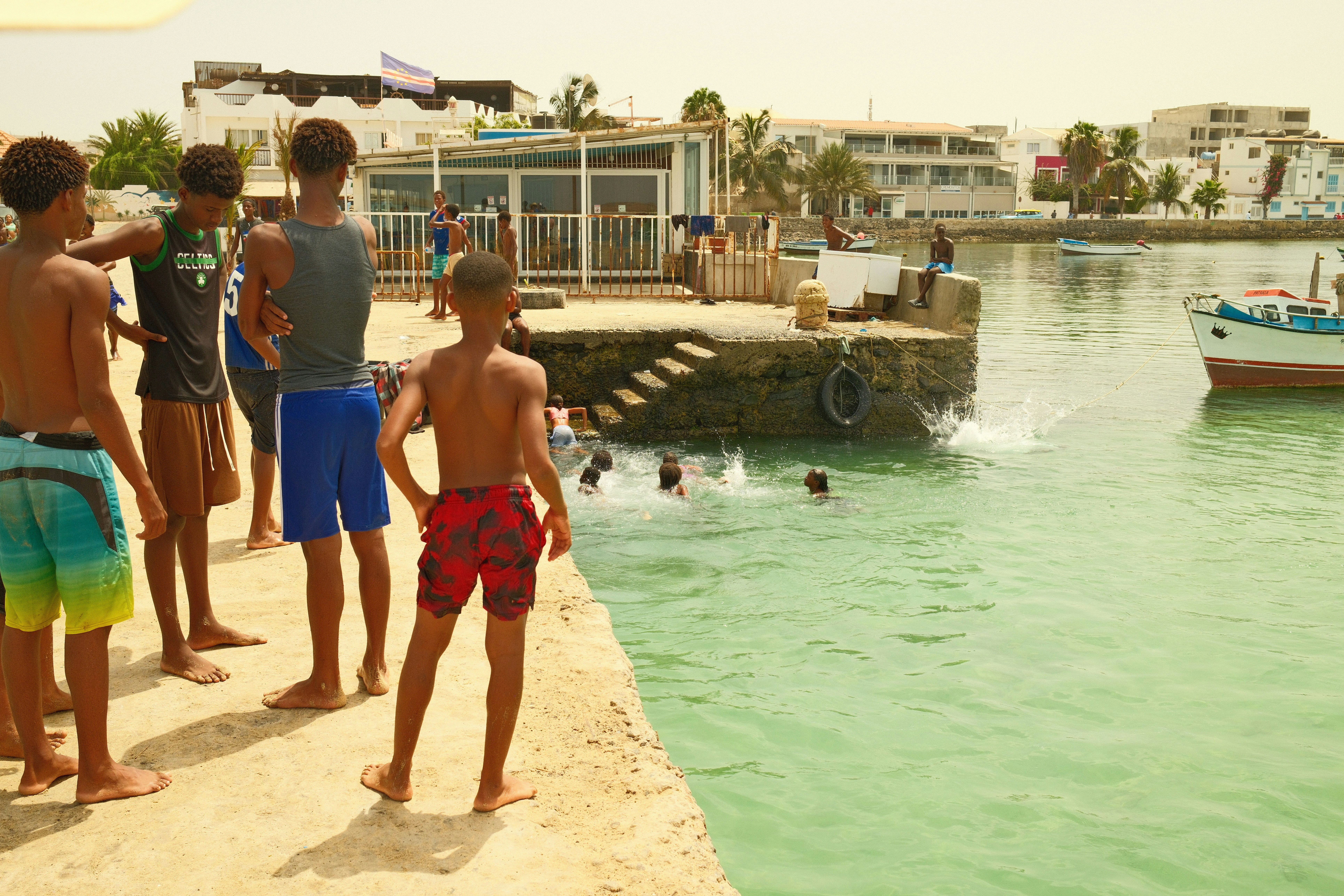 Children gather at a sunlit harbor edge, peering into turquoise water near a dock