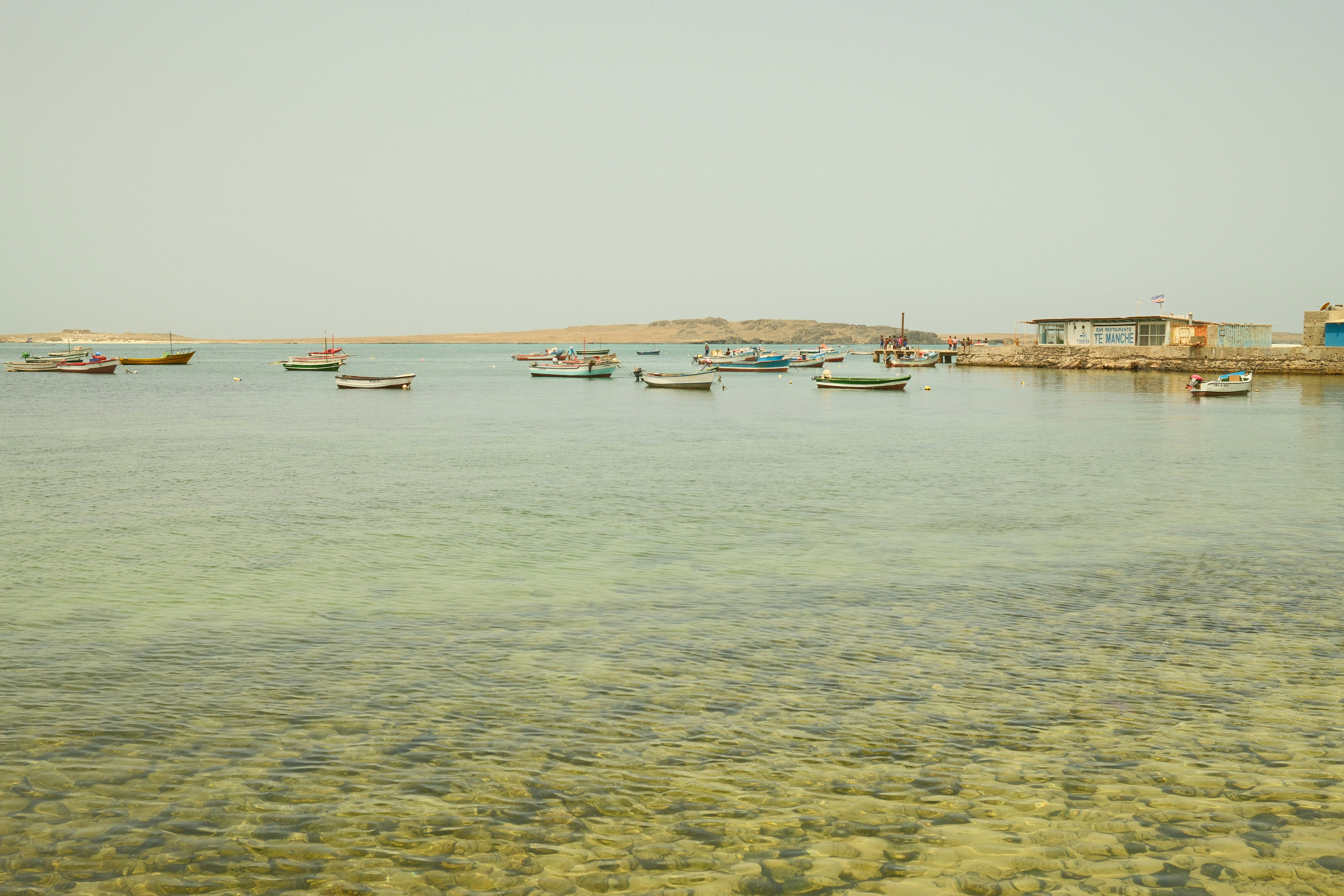 Calm coastal scene with a line of small boats anchored near a shallow, rocky shoreline and a distant hut on a sandy spit.
