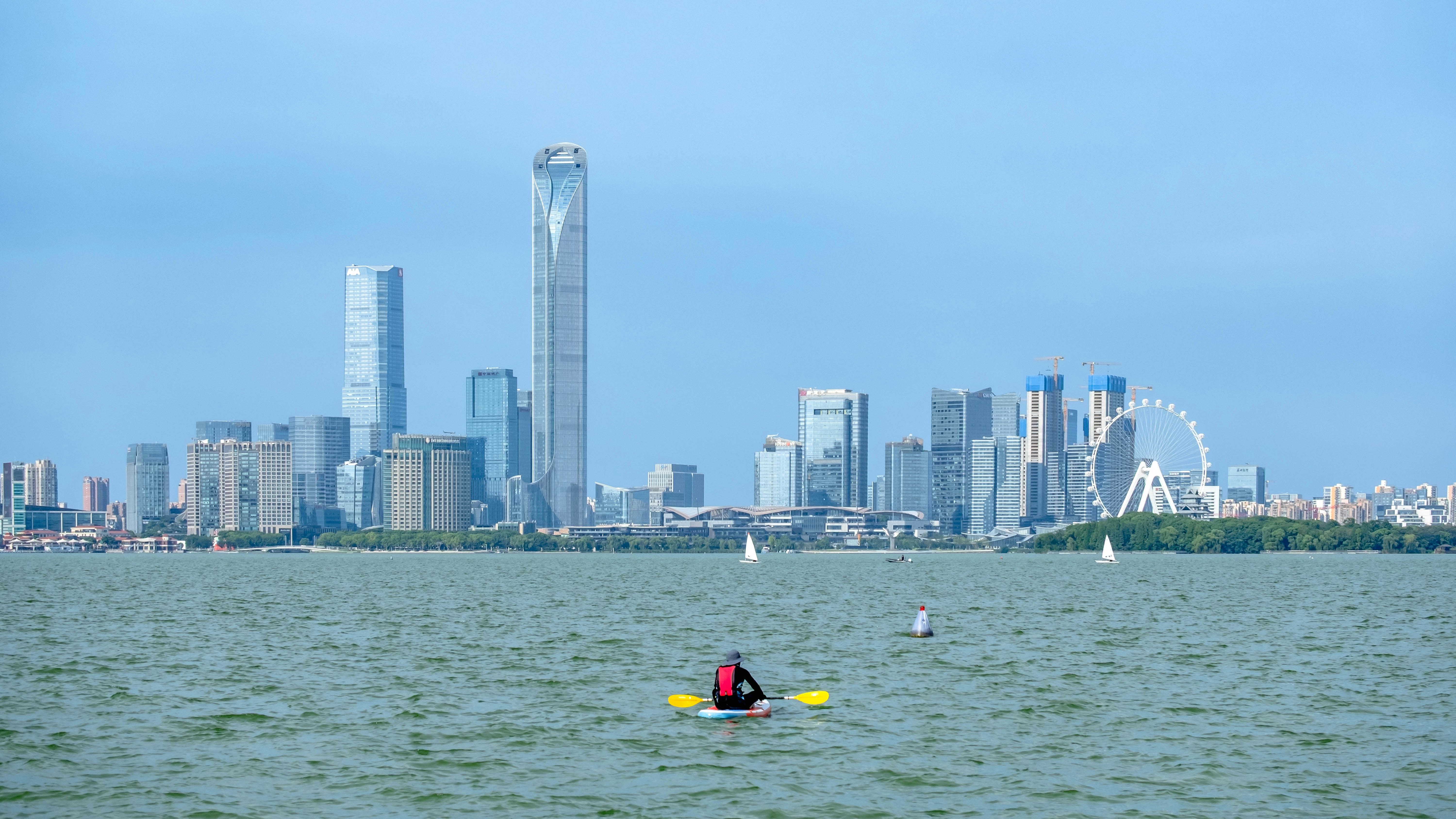 Kayaker paddles on greenish water with a modern skyline and a white Ferris wheel in the distance.