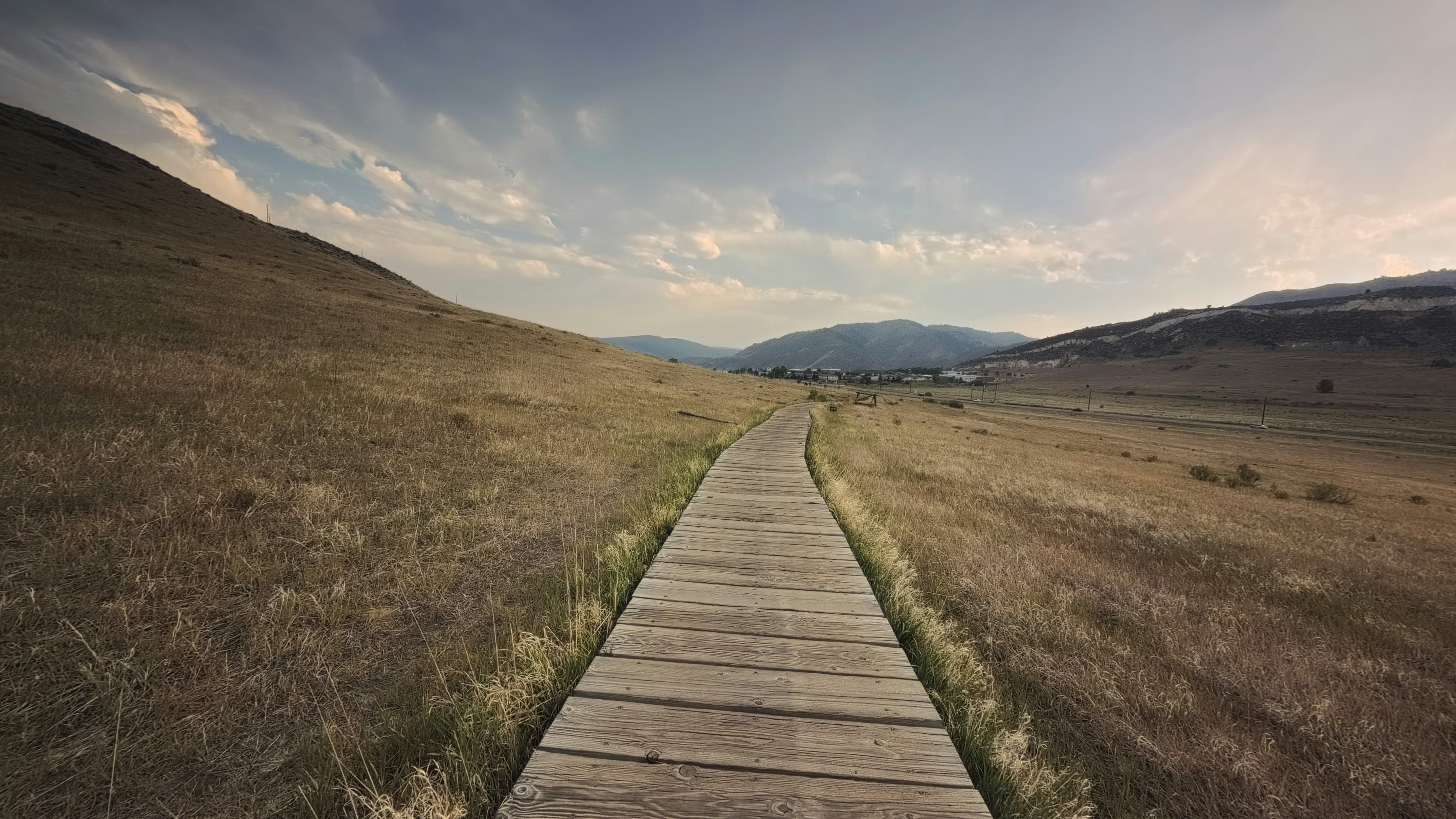 A wooden boardwalk cuts through a dry, golden hillside toward distant mountains under a soft sky. The scene emphasizes path and horizon with a serene, expansive landscape.