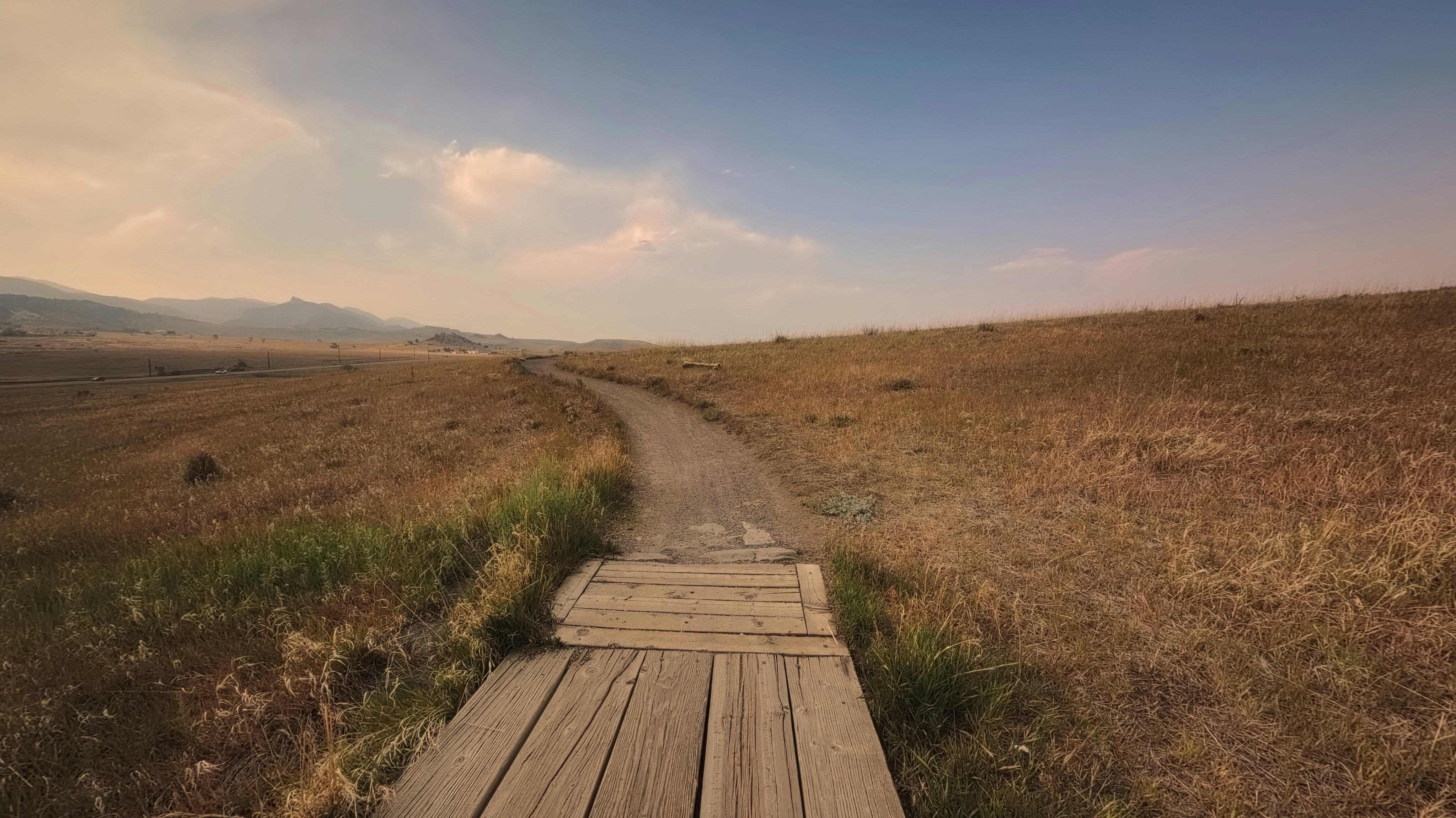 Rounded wooden boardwalk leading through sunlit prairie toward distant hills