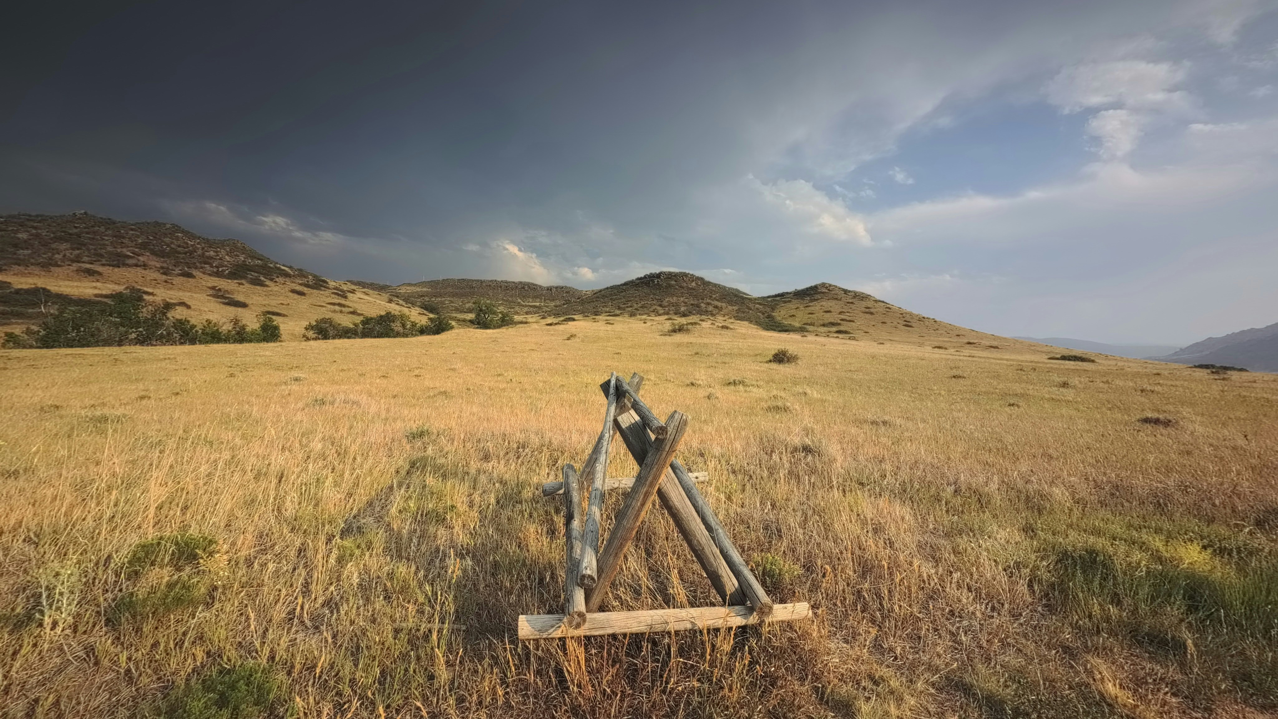 Golden grassland with a rustic wooden tripod in the foreground under a dramatic sky; a wide landscape emphasizing open space and distant hills.