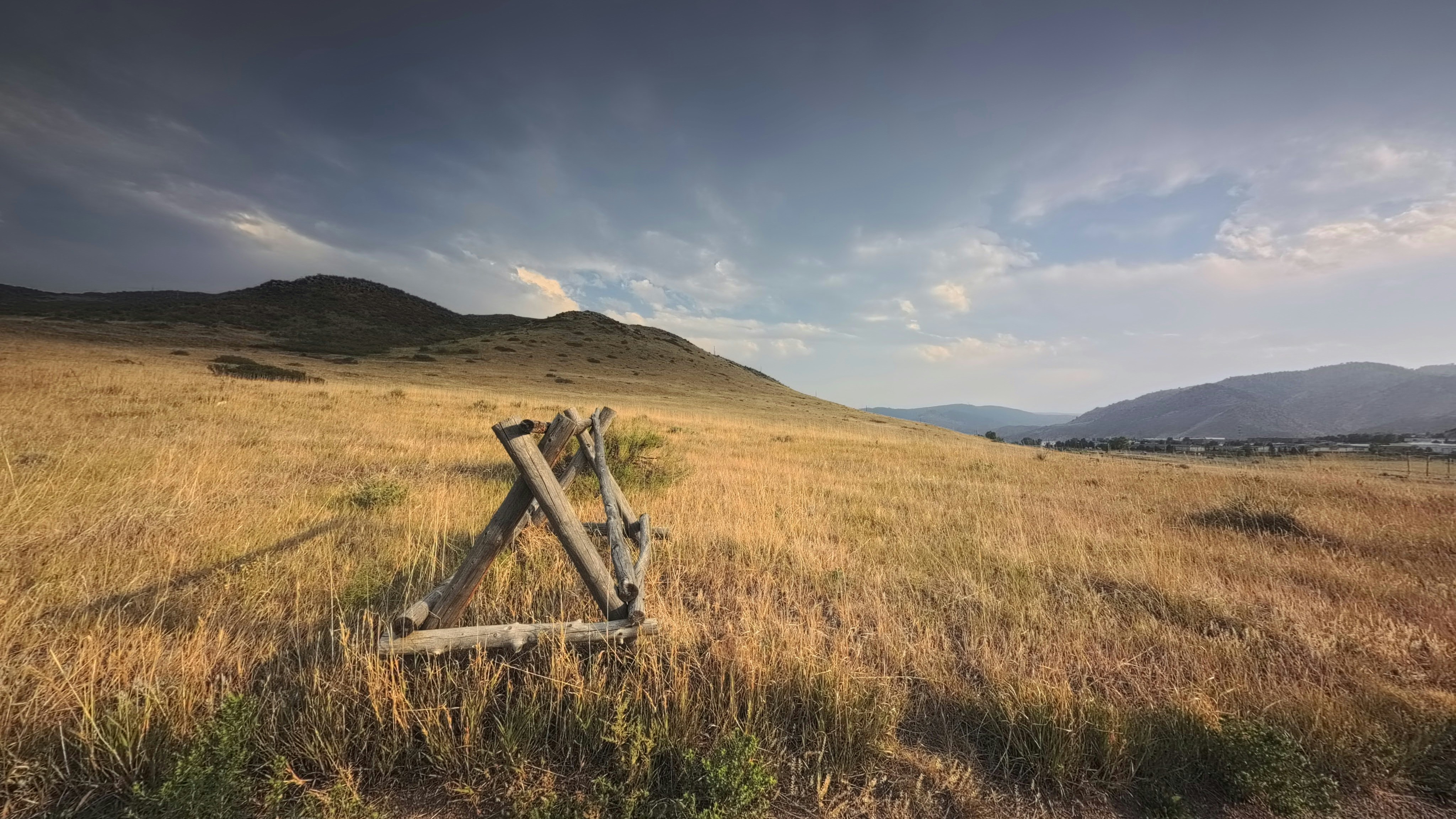 Golden grass field with a weathered wooden fence in the foreground, rolling hills in the distance under a dramatic cloudy sky.