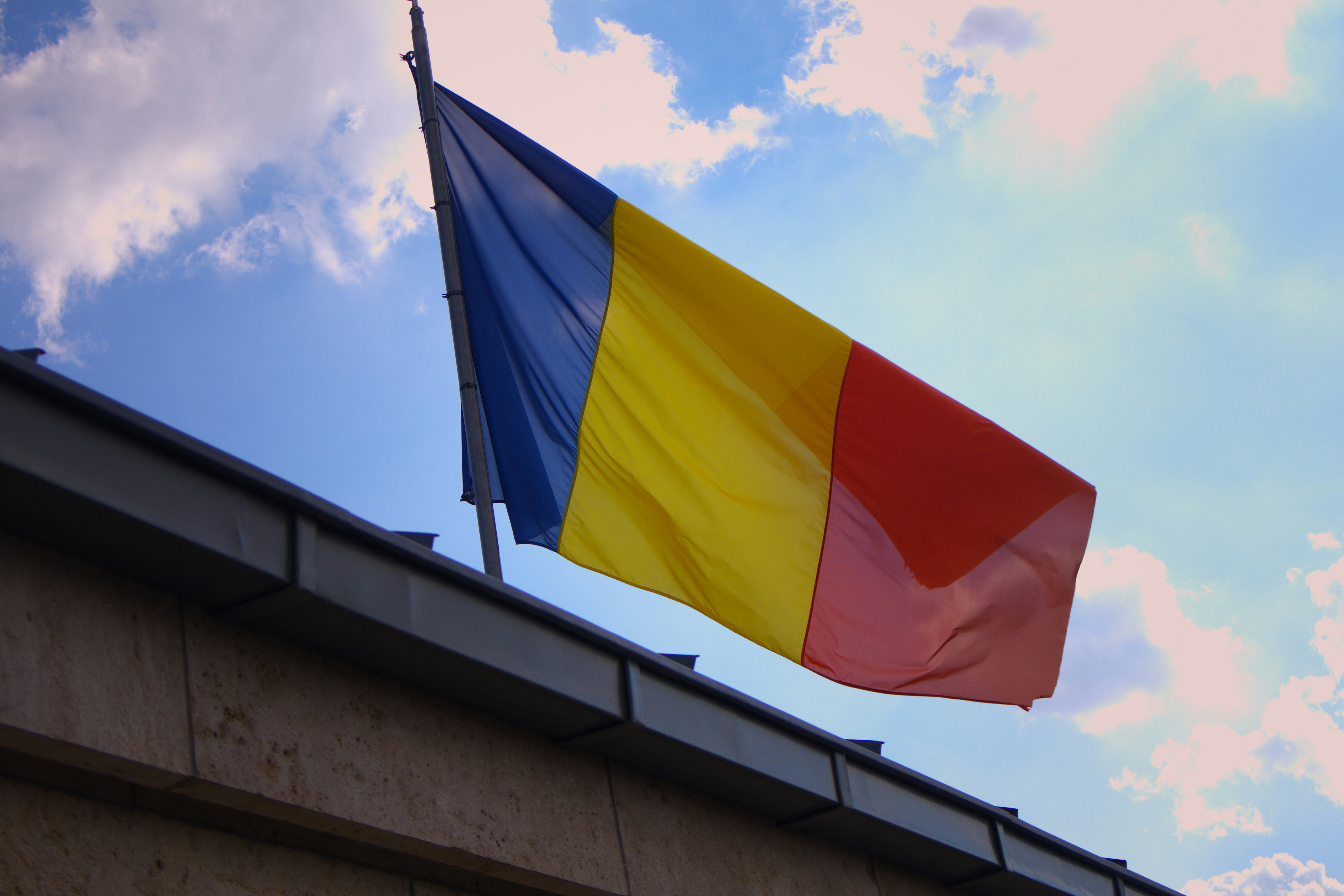 A tri-color flag (blue, yellow, red) waves above a building against a bright sky, emphasizing motion and wind.