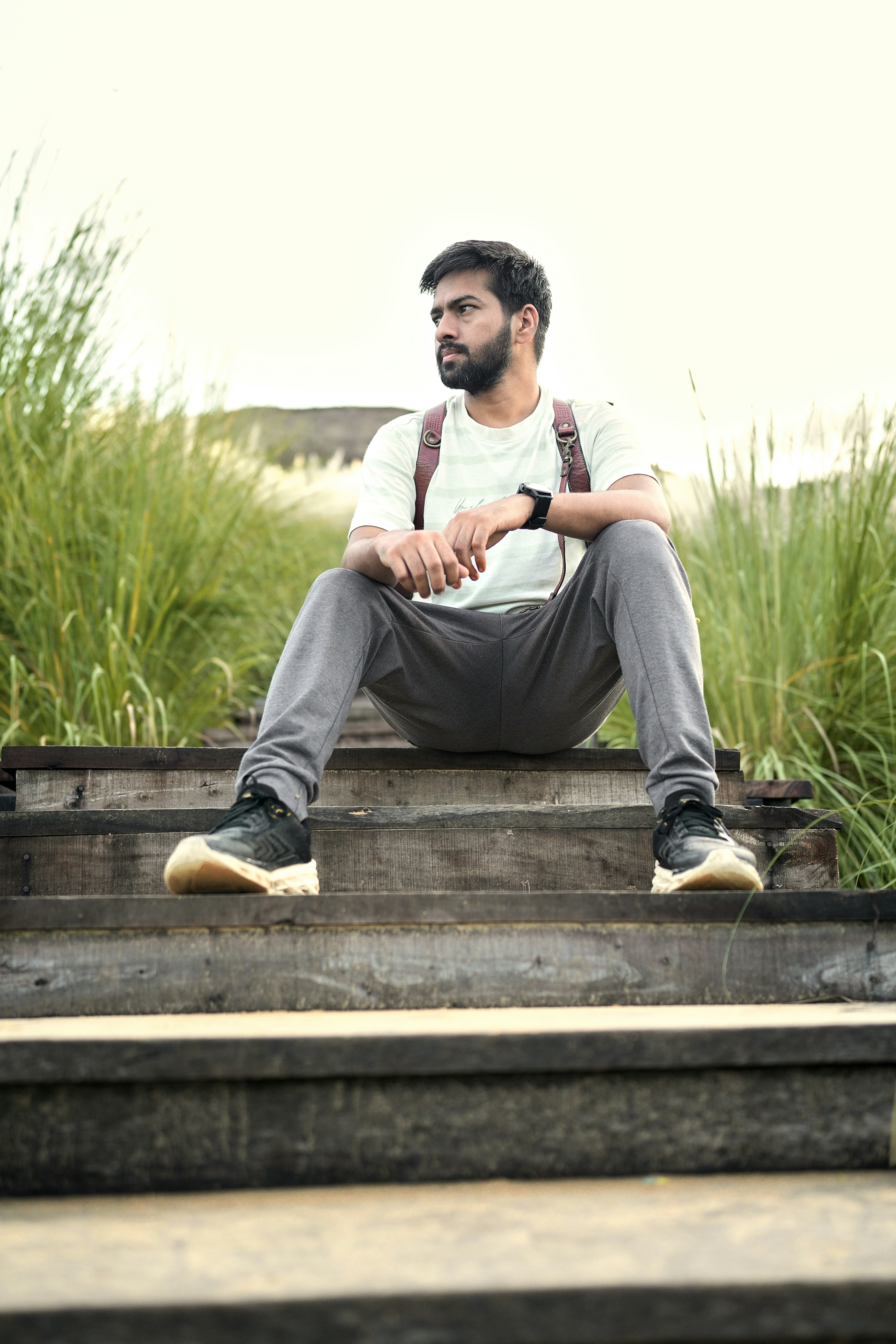 Man seated on weathered wooden steps among tall reeds, wearing a white shirt and suspenders, gazing off to the right; a calm outdoor portrait with natural backlight. Logo-like watch on wrist.