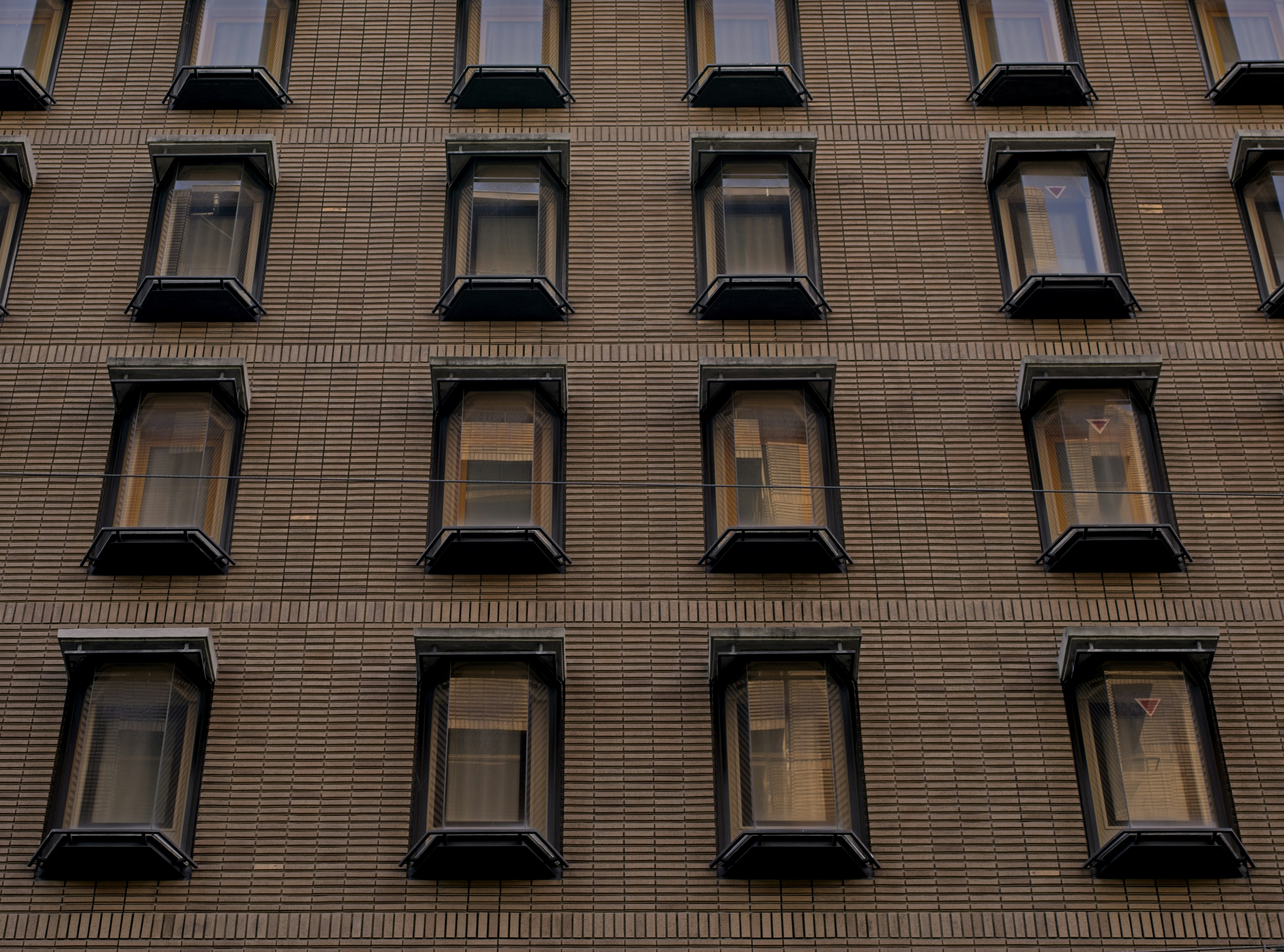 A striking grid of projecting bay windows repeats across a warm brown brick facade, each framed in black metal with slight variations in curtains, blinds, and small red triangle stickers visible through the glass. The rhythmic symmetry and textured brickwork create a graphic, almost abstract composition typical of Osaka's dense urban architecture. Found just off the bustling streets of the city's trendy district, this quiet architectural detail offers a contrasting, contemplative pause from the vibrant storefronts and street art nearby on a hot July afternoon.