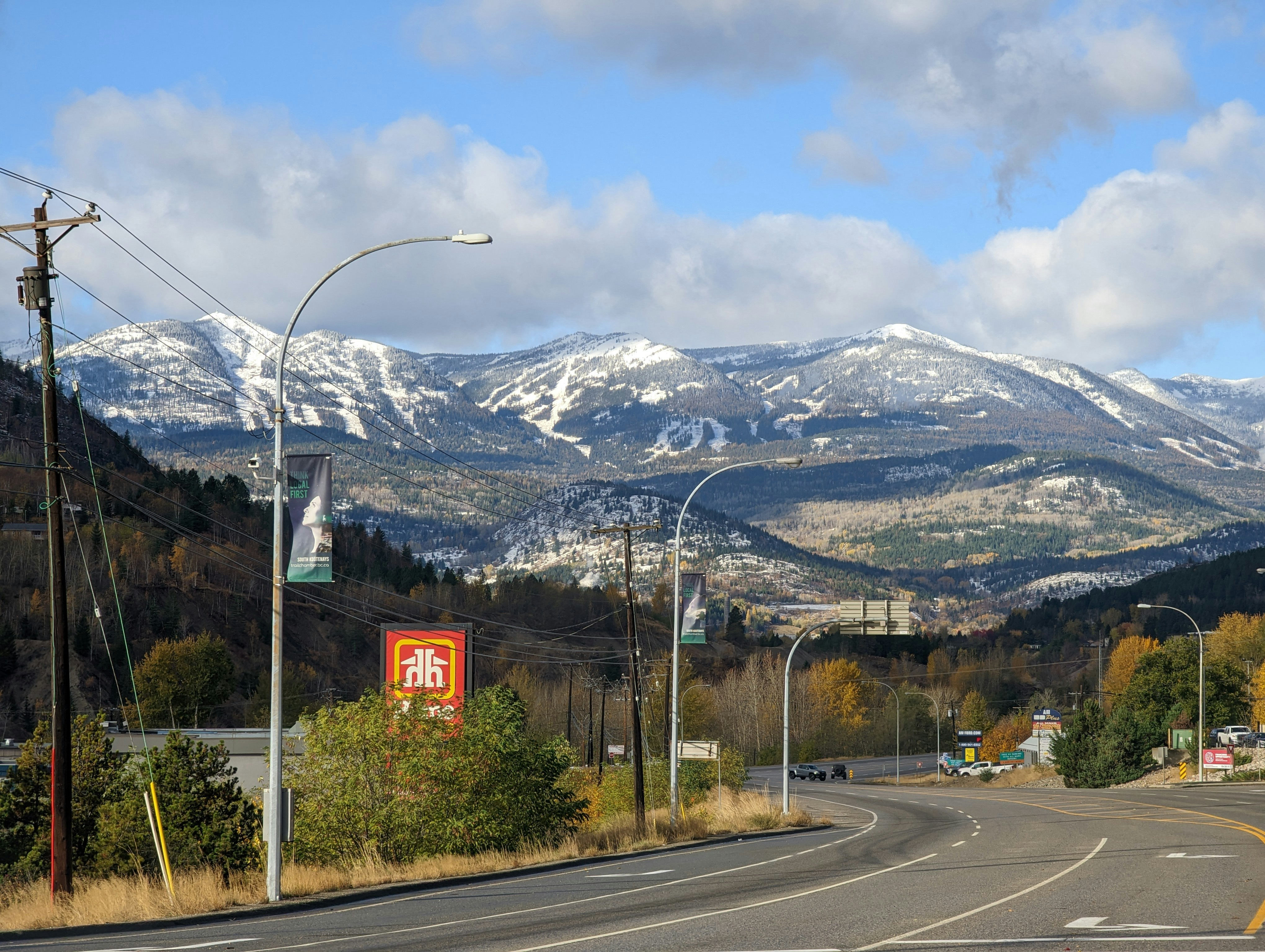 Wide mountain highway with snow-capped peaks in the distance and a roadside scene featuring utility poles and a red sign, conveying a calm, rural commute.