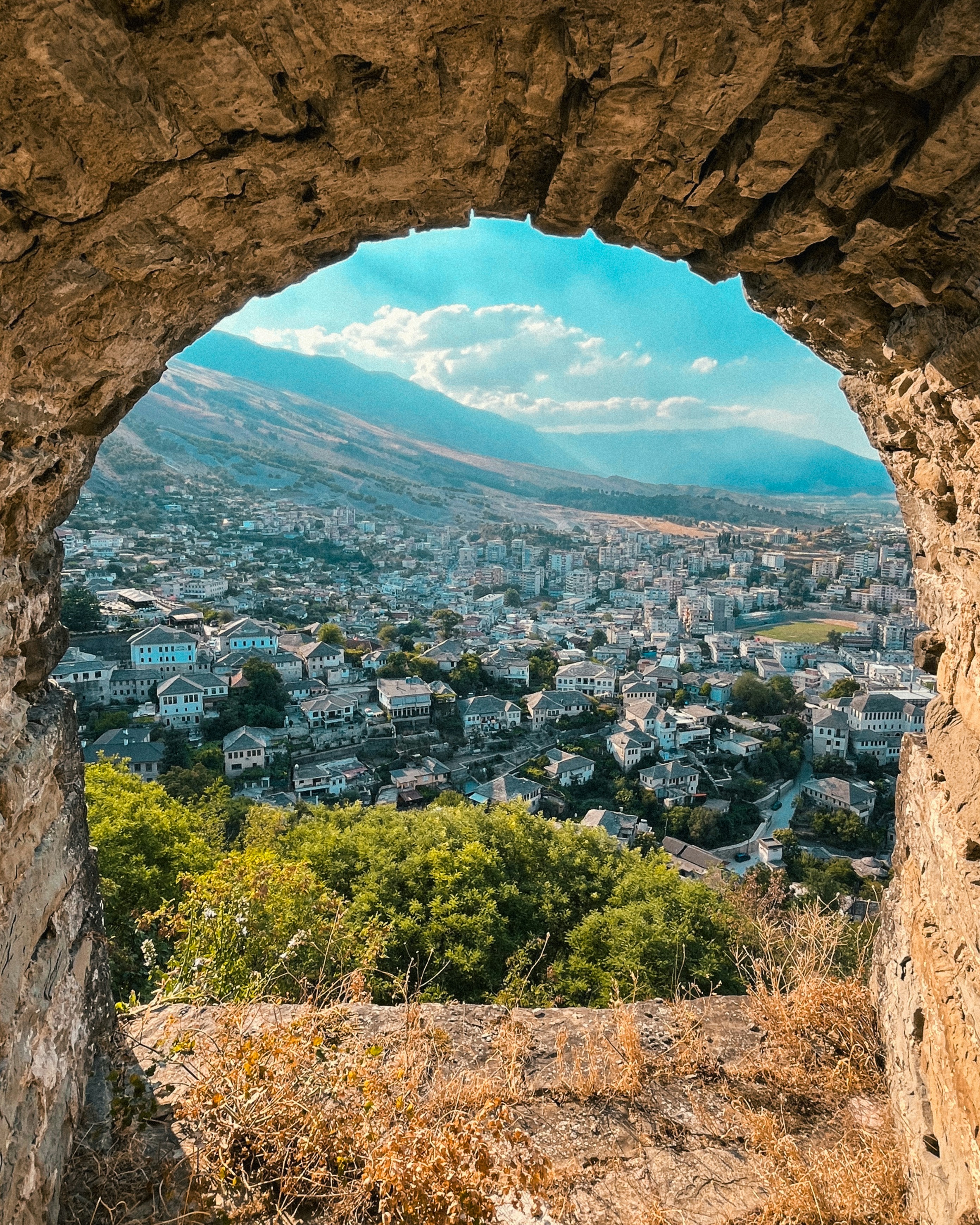 A valley city viewed through a rugged stone arch, with rooftops and distant mountains under a bright sky.