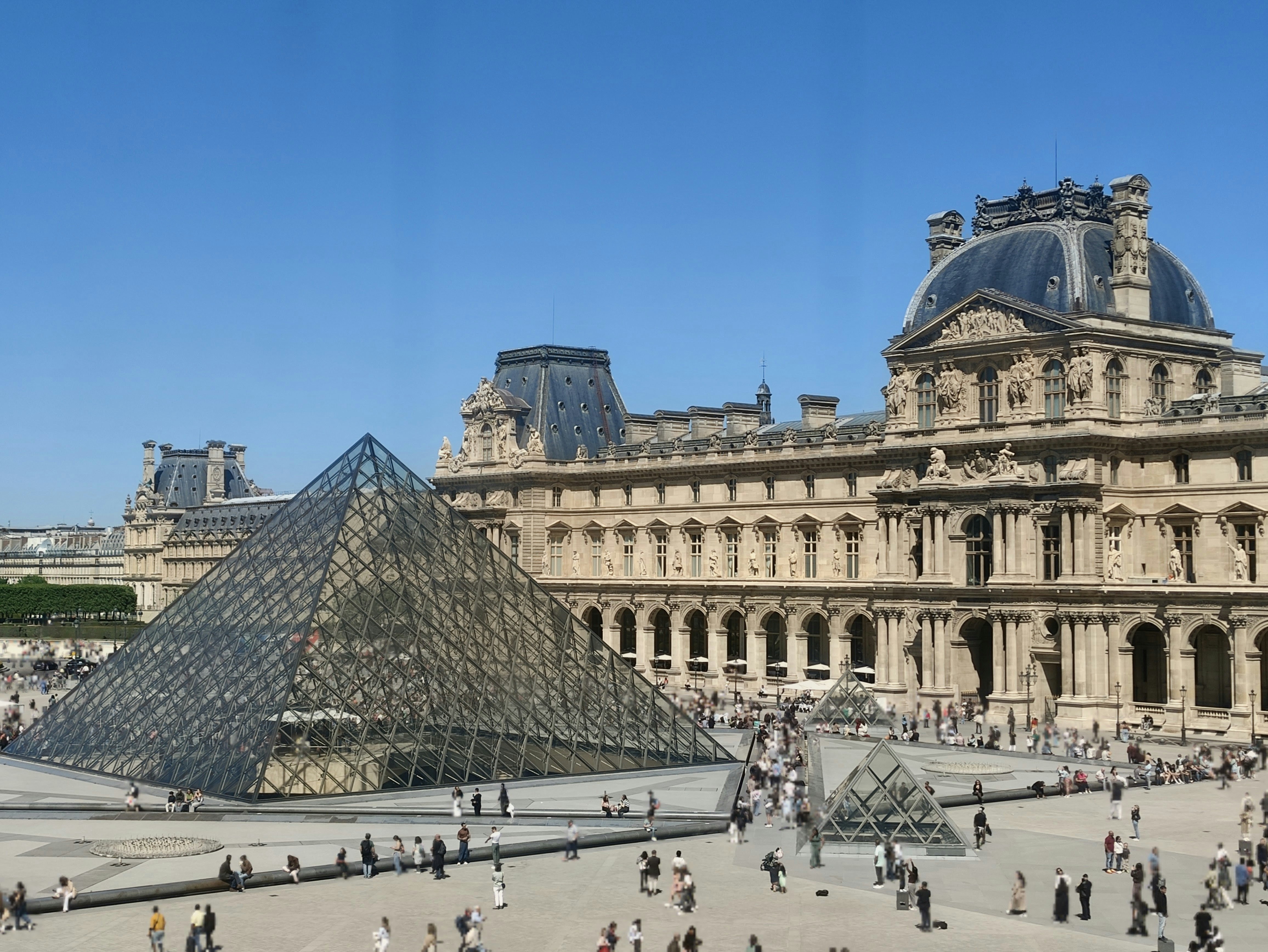 Sunlit Louvre courtyard with the glass pyramids and historic façade, crowds dispersed around the iconic architecture. Photograph captures the blend of modern glass geometry and classical stone detailing.