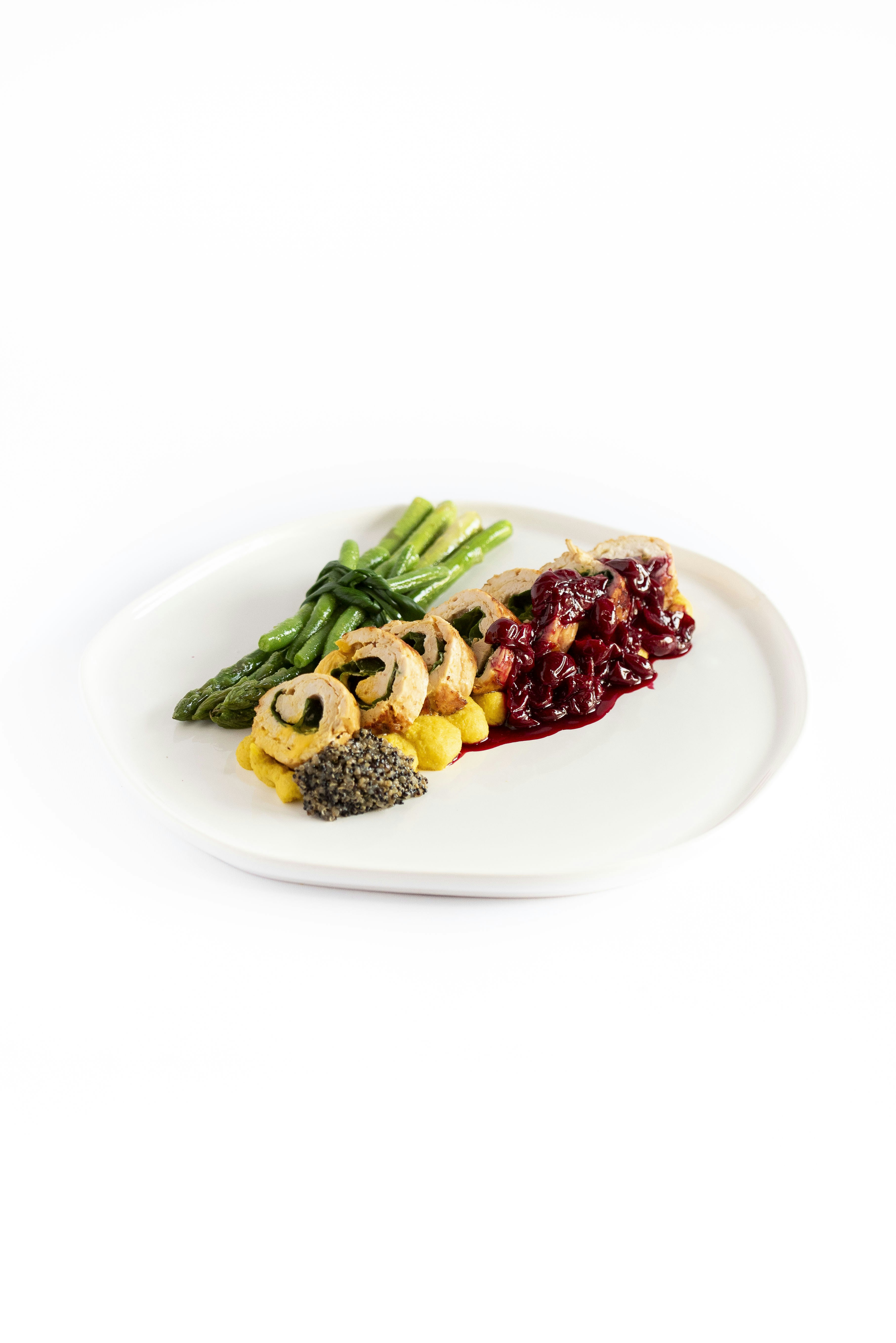 Culinary plate featuring rolled protein, yellow dollops, green asparagus, and a cranberry sauce on a white dish.