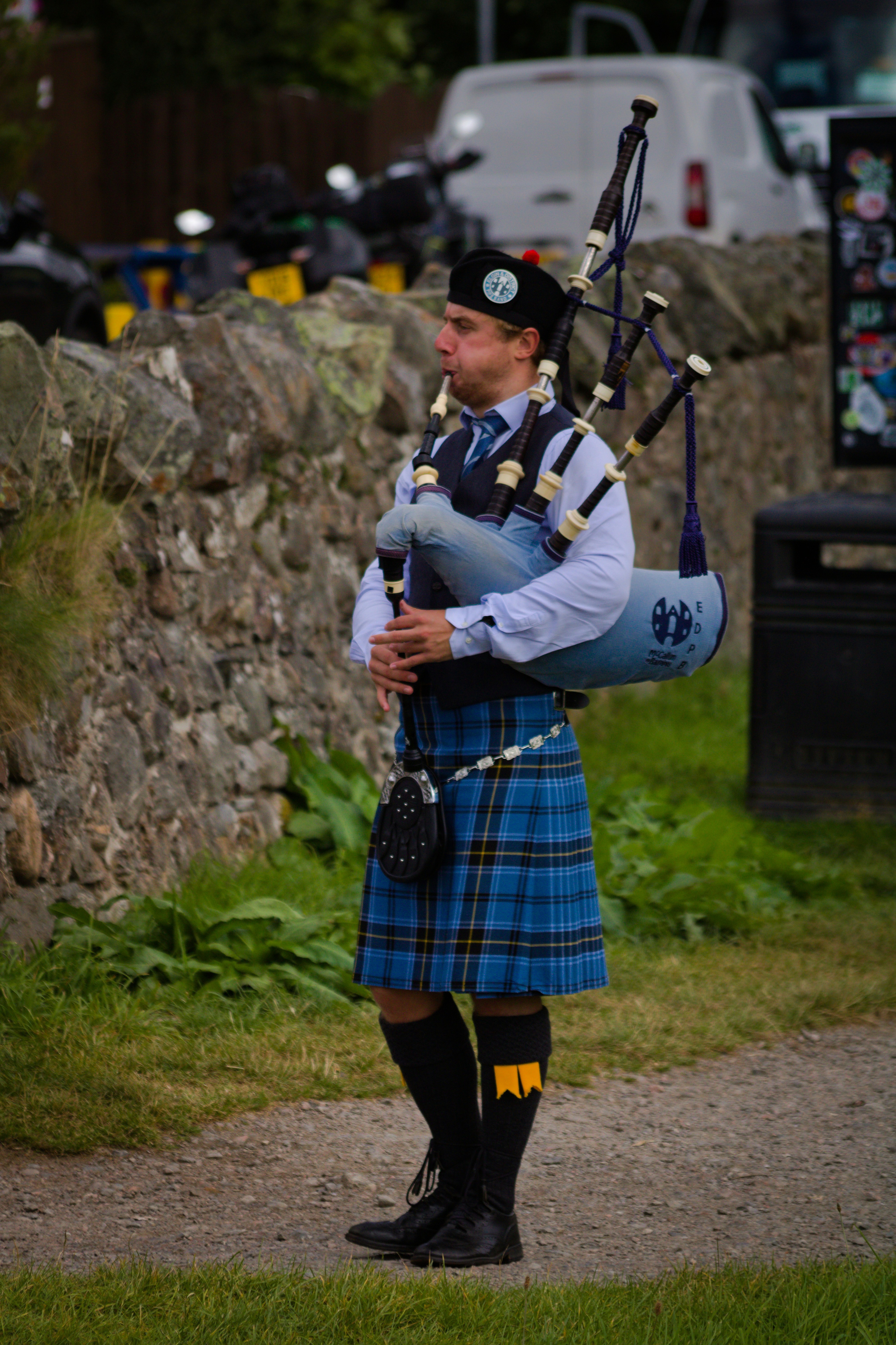 Highland Piper in a Blue Plaid kilt Plays the Bagpipes by a Stone Wall