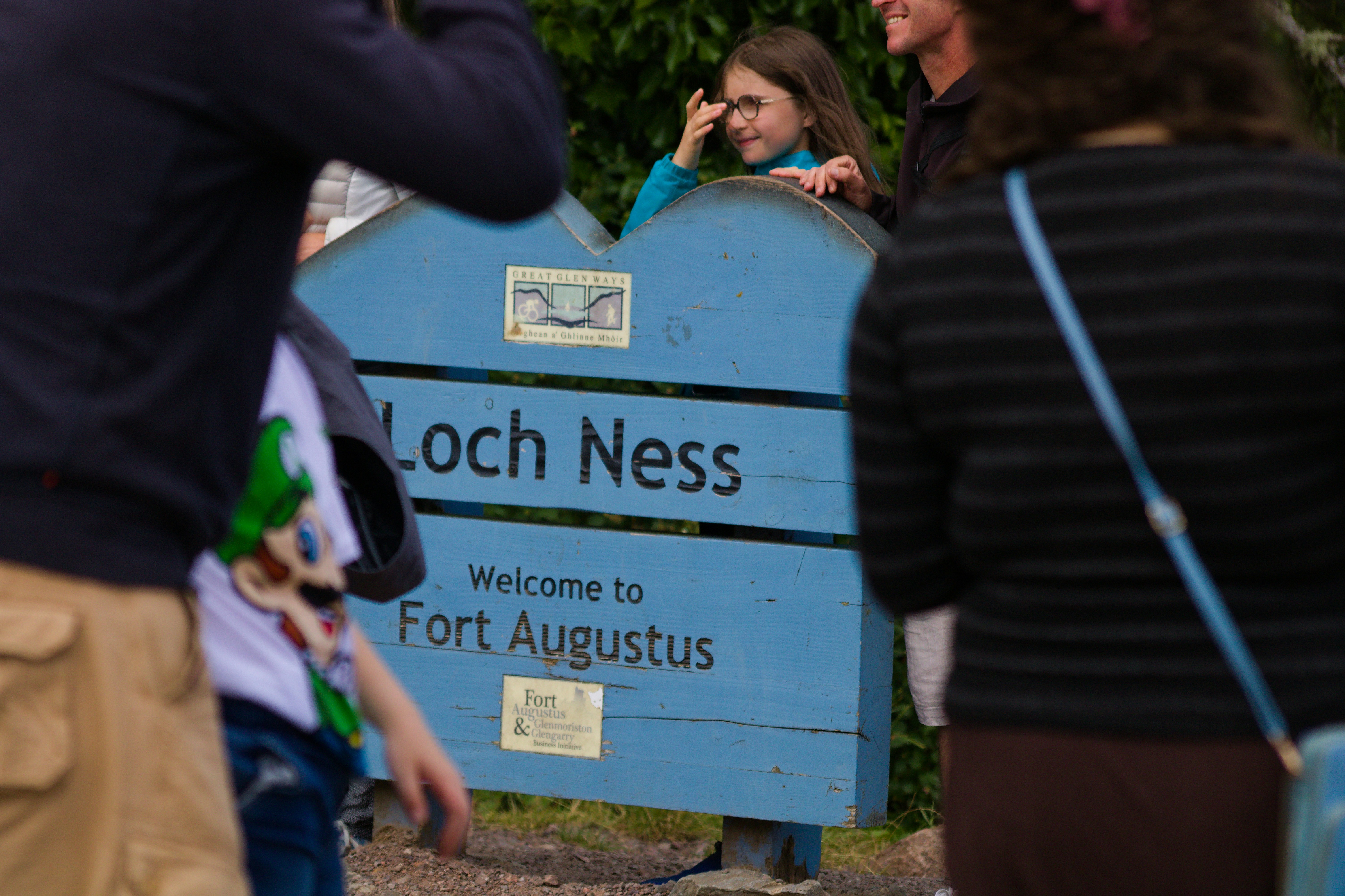 Blue Welcome Sign at Fort Augustin Cutout: People Pausing by a Child Behind a Park Barrier