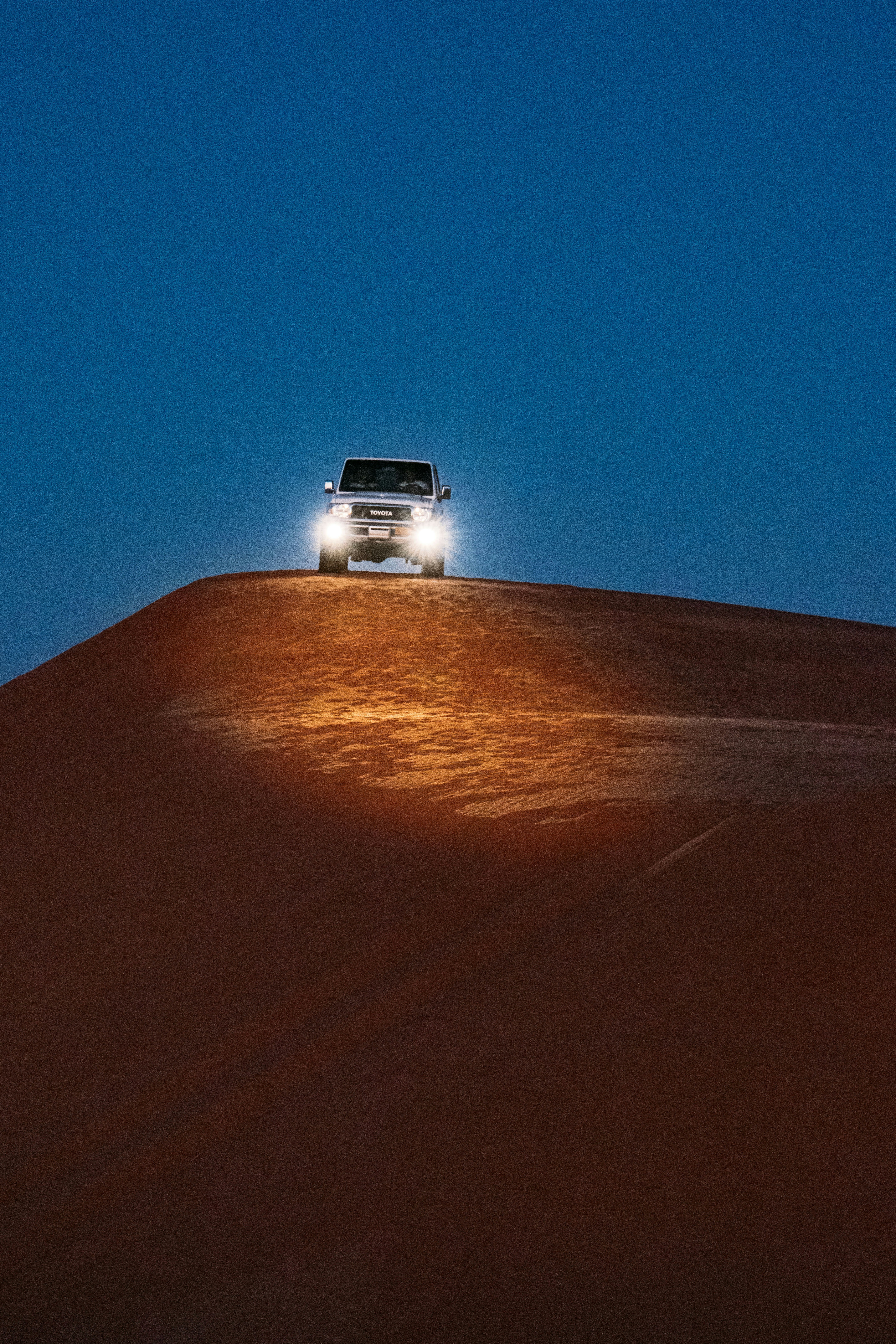A white SUV sits with its headlights on atop a reddish dune against a darkening blue sky, emphasizing isolation and a quiet moment at the crest. (Logo/vehicle present).