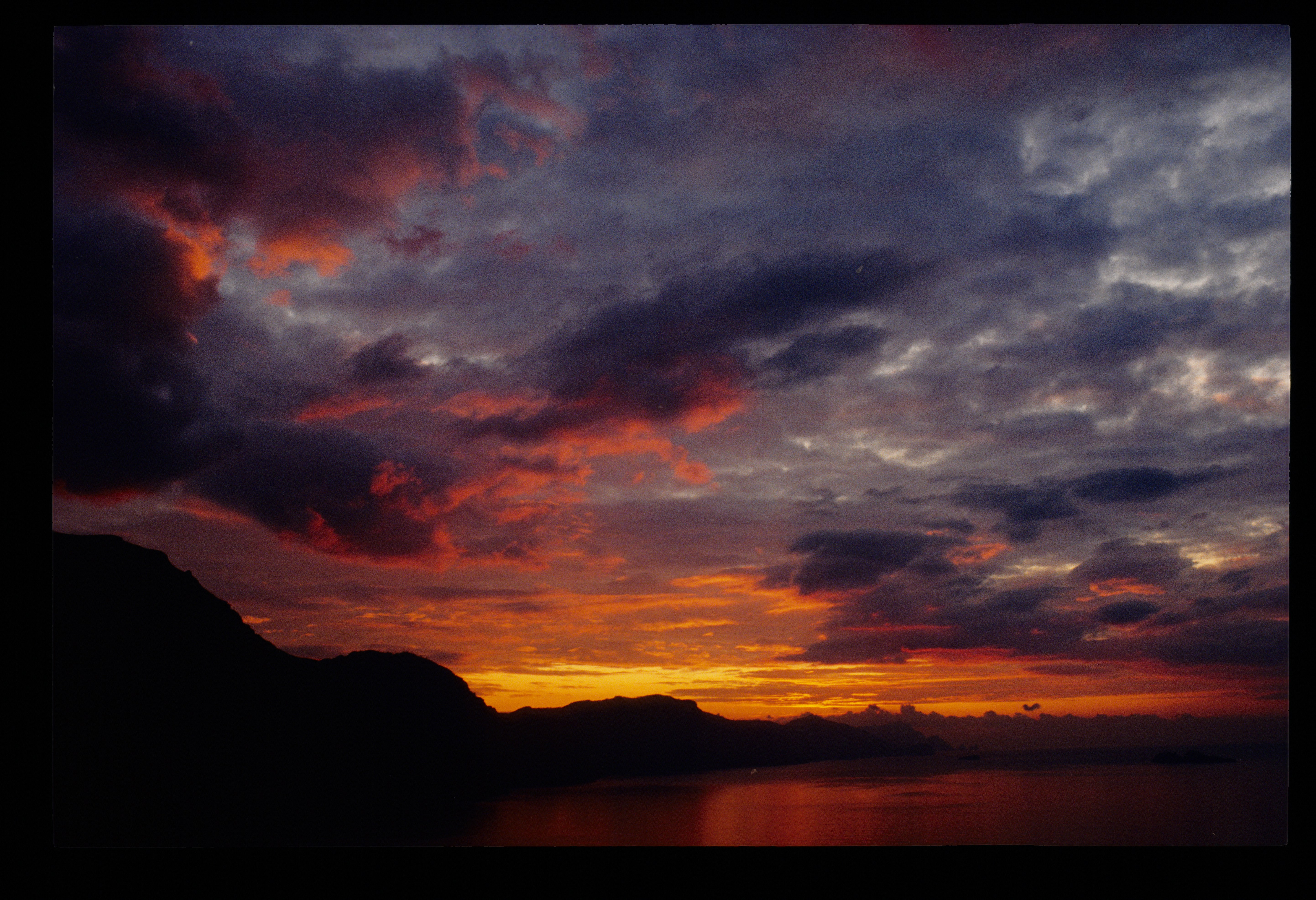 Soft-edged silhouettes of a coastal landscape at sunset with vibrant orange and purple sky over calm water. A tranquil, wide scene focusing on the horizon glow.