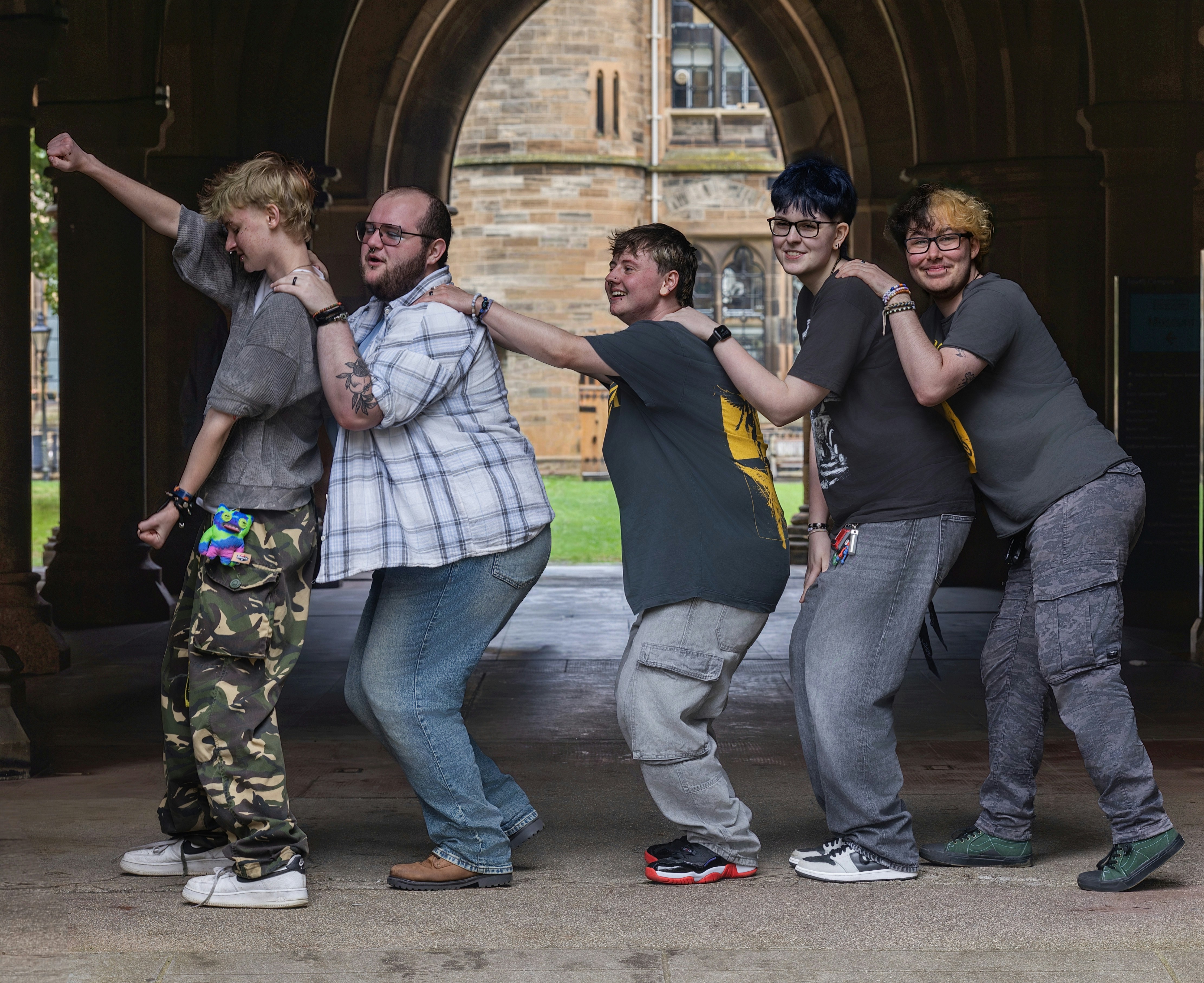 A group of five friends in a staged conga line under an arched stone walkway, each linking arms and smiling as they pose toward the camera. The scene emphasizes camaraderie and casual outdoor style.