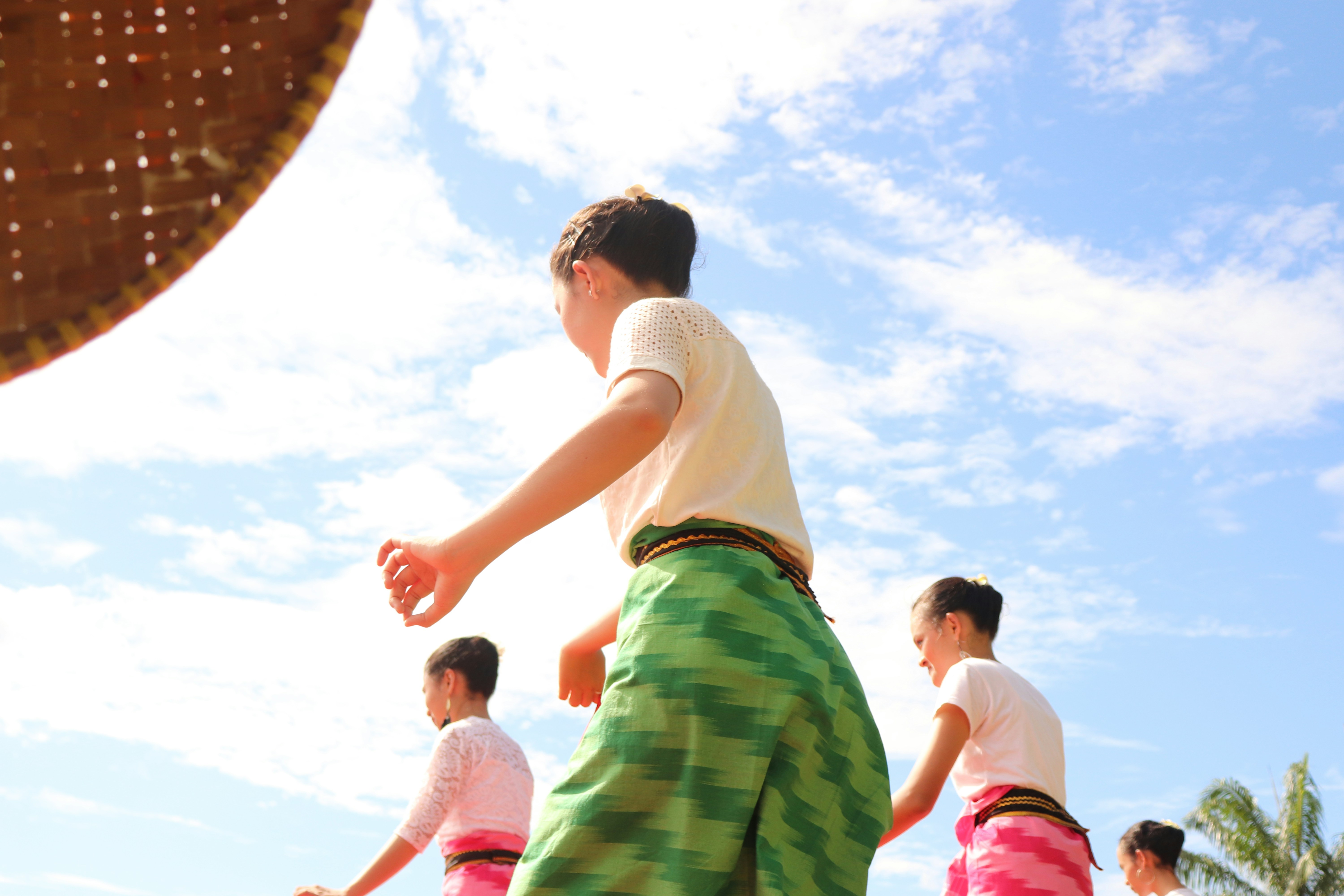 Brightly dressed children walk toward the edge of the frame against a blue sky and wispy clouds; a portion of a shaded structure is visible at the top left. This scene captures a carefree outdoor moment with a shallow perspective.