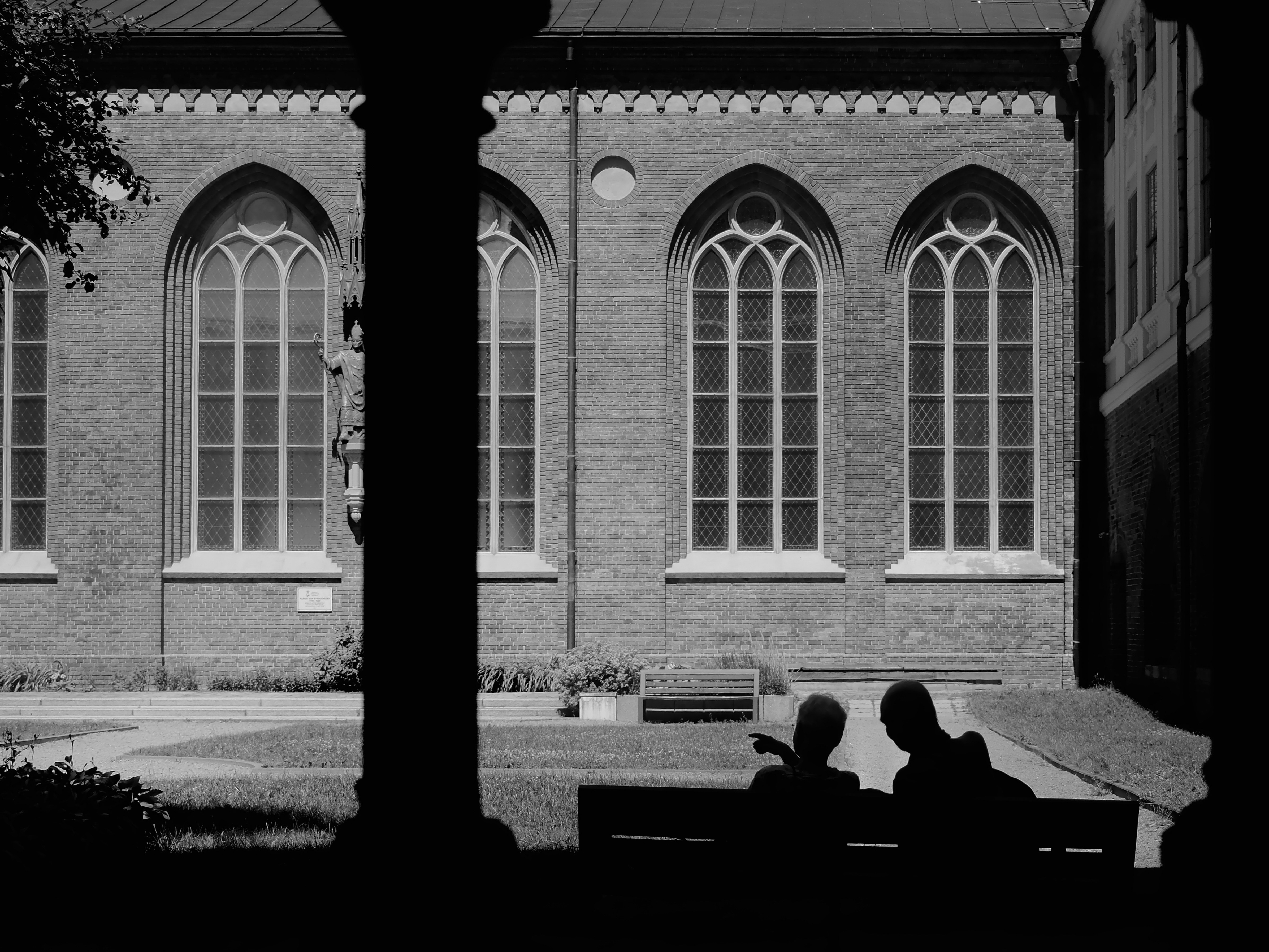 High-contrast grayscale scene of a church courtyard with large arched windows; three figures seated on a bench in shadow beneath a column, with brick walls and trimmed lawn in the background.