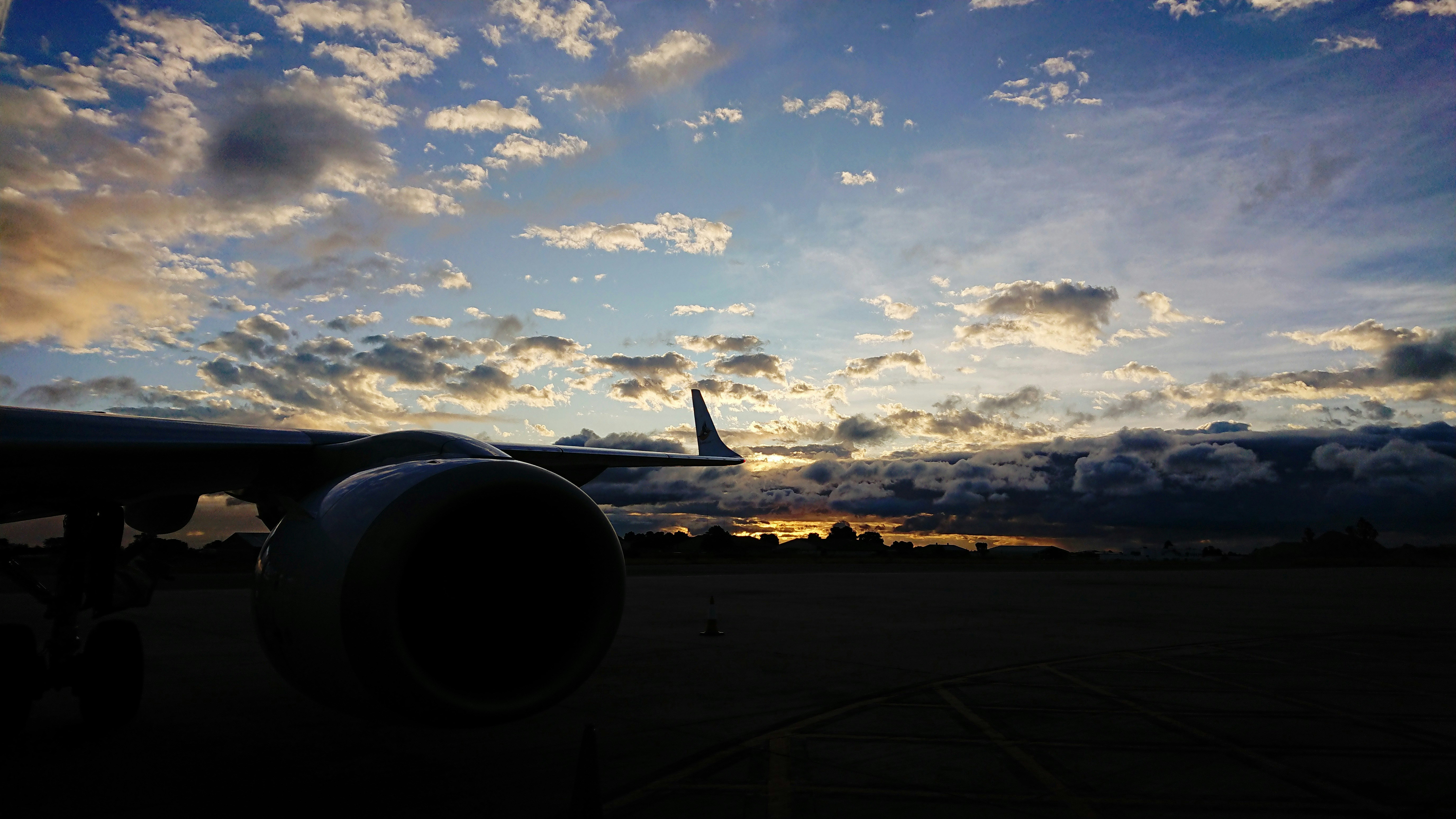 Dark silhouette of an airplane on tarmac at sunset, engine nacelle and wing visible in foreground with dramatic sky and clouds behind.