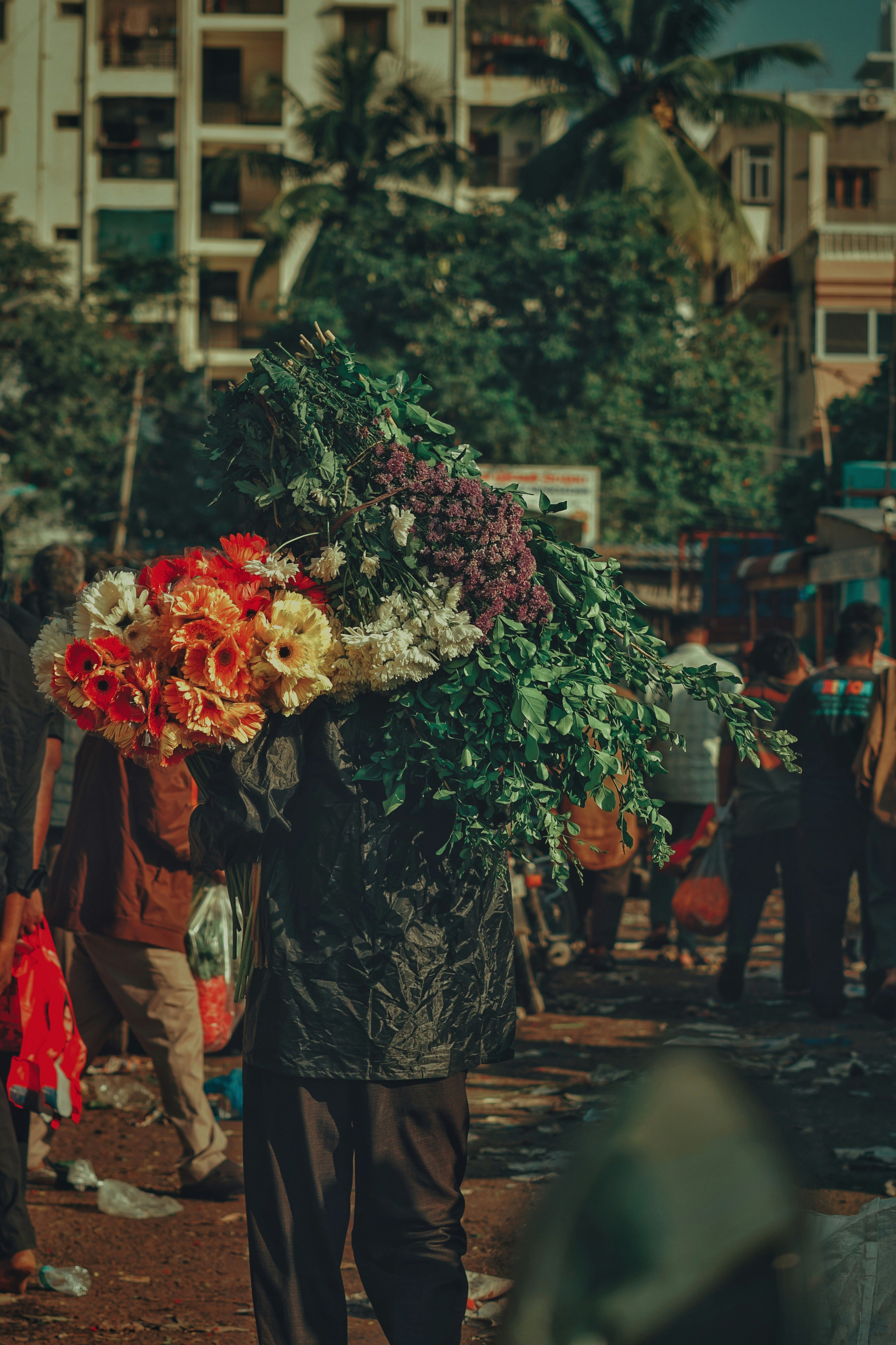 Vibrant flower arrangement carried by a vendor in a crowded urban market, with buildings and people in the background.