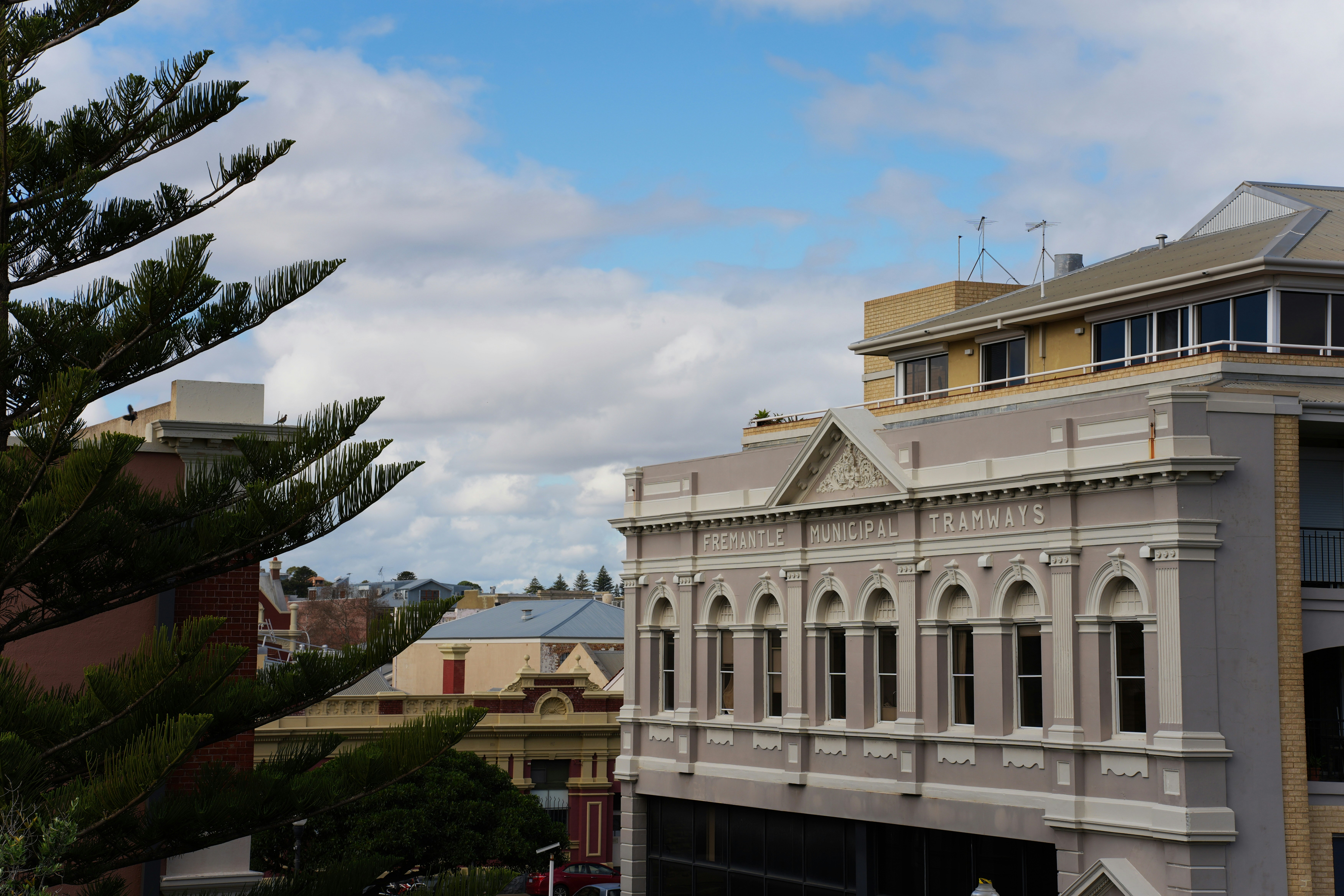 Historic-looking building with arched windows dominates the frame as a tree and other structures sit to the left; a bright daytime sky serves as a backdrop for urban architecture.