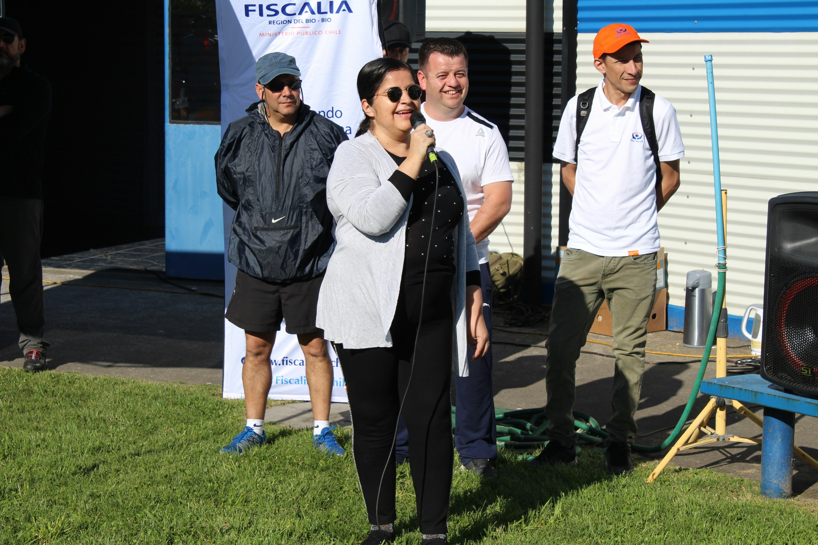 Speaker in light gray cardigan addresses a small crowd outdoors