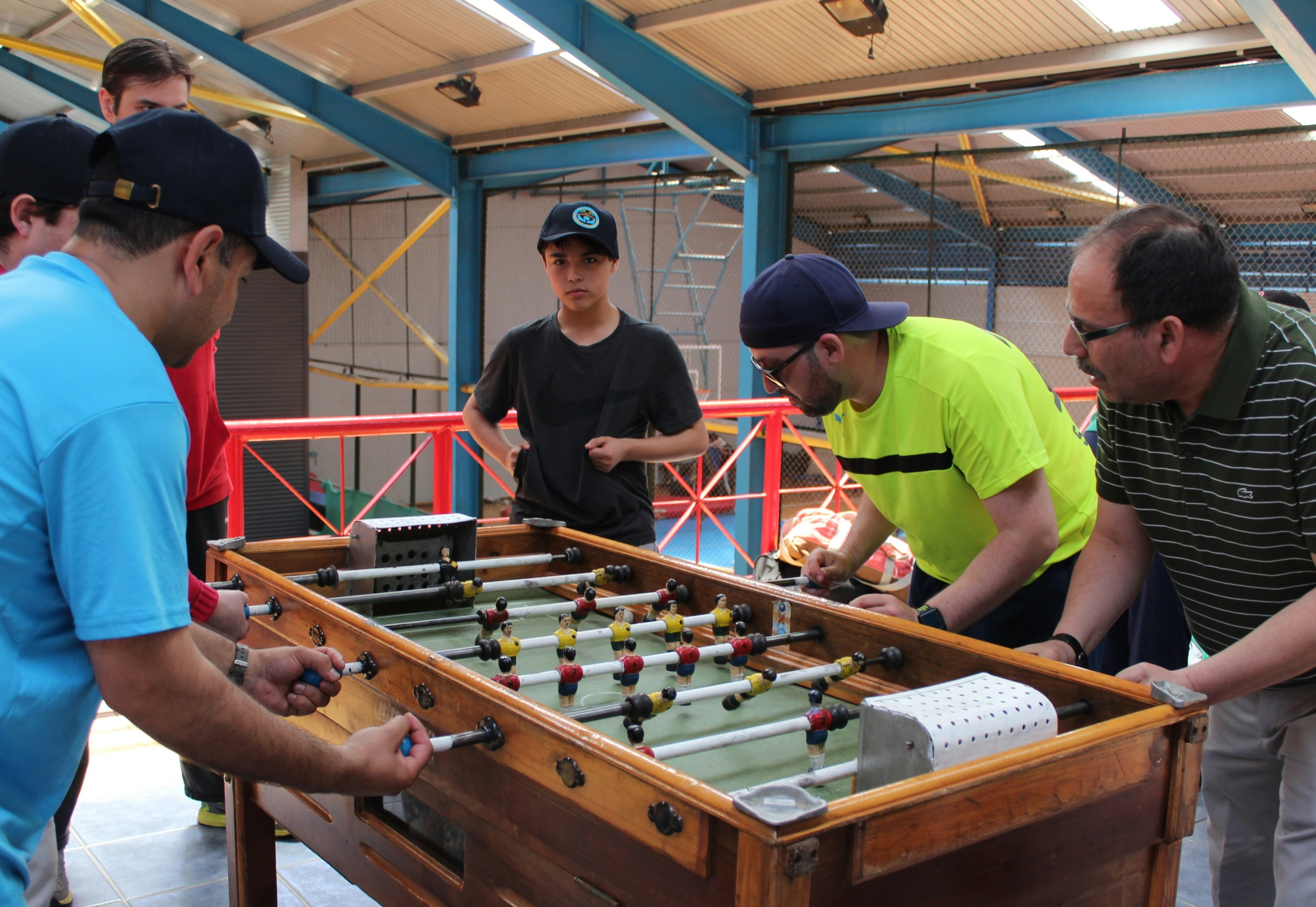 A group of players gathers around a foosball table during a casual match in a bright indoor space. The scene centers on the active players and the table controls, conveying teamwork and competition.