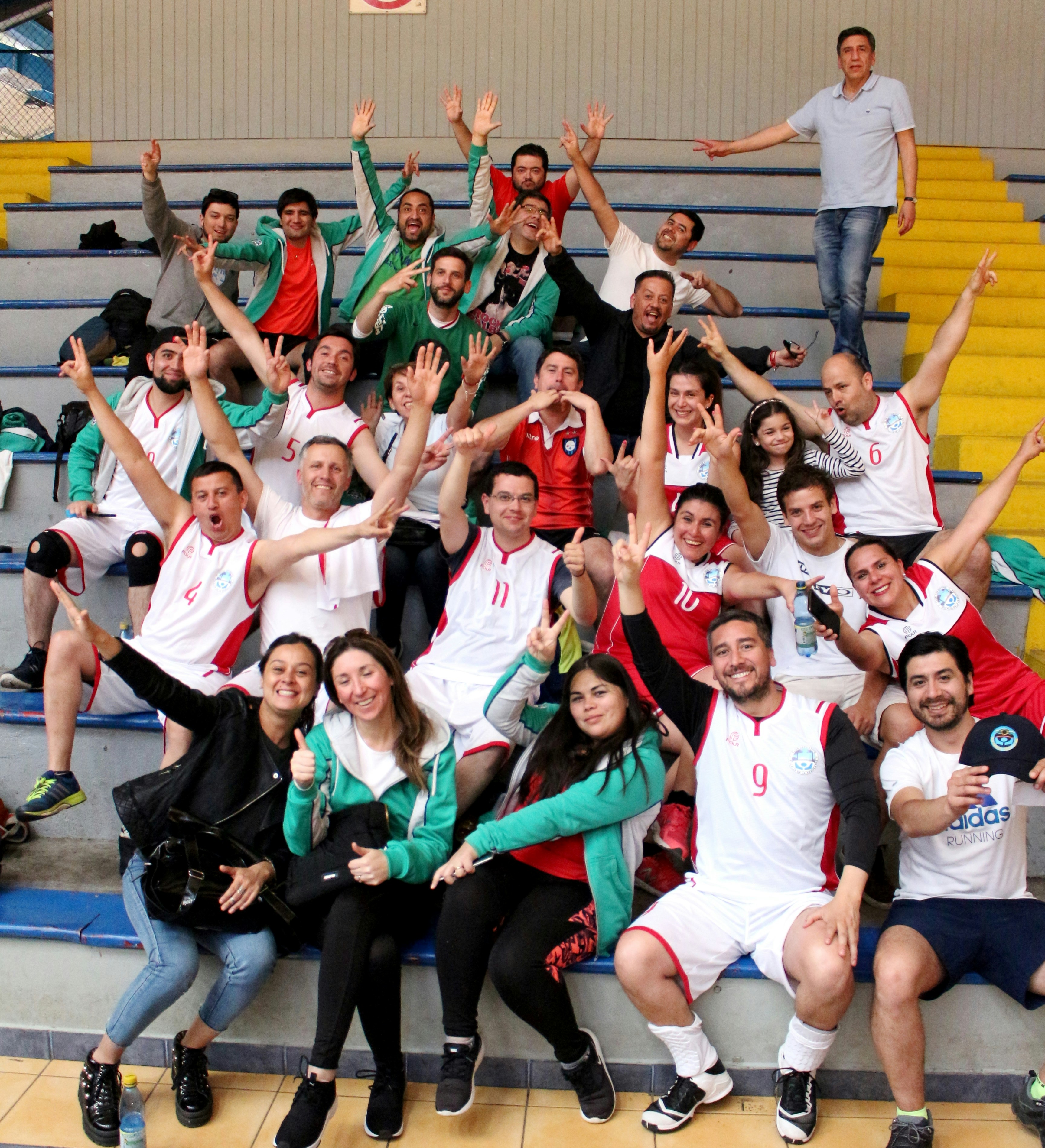 Group of athletes in white and red uniforms posing on bleachers, arms raised in celebration; crowd includes coaches and supporters, energy-filled team photo in a sports hall.
