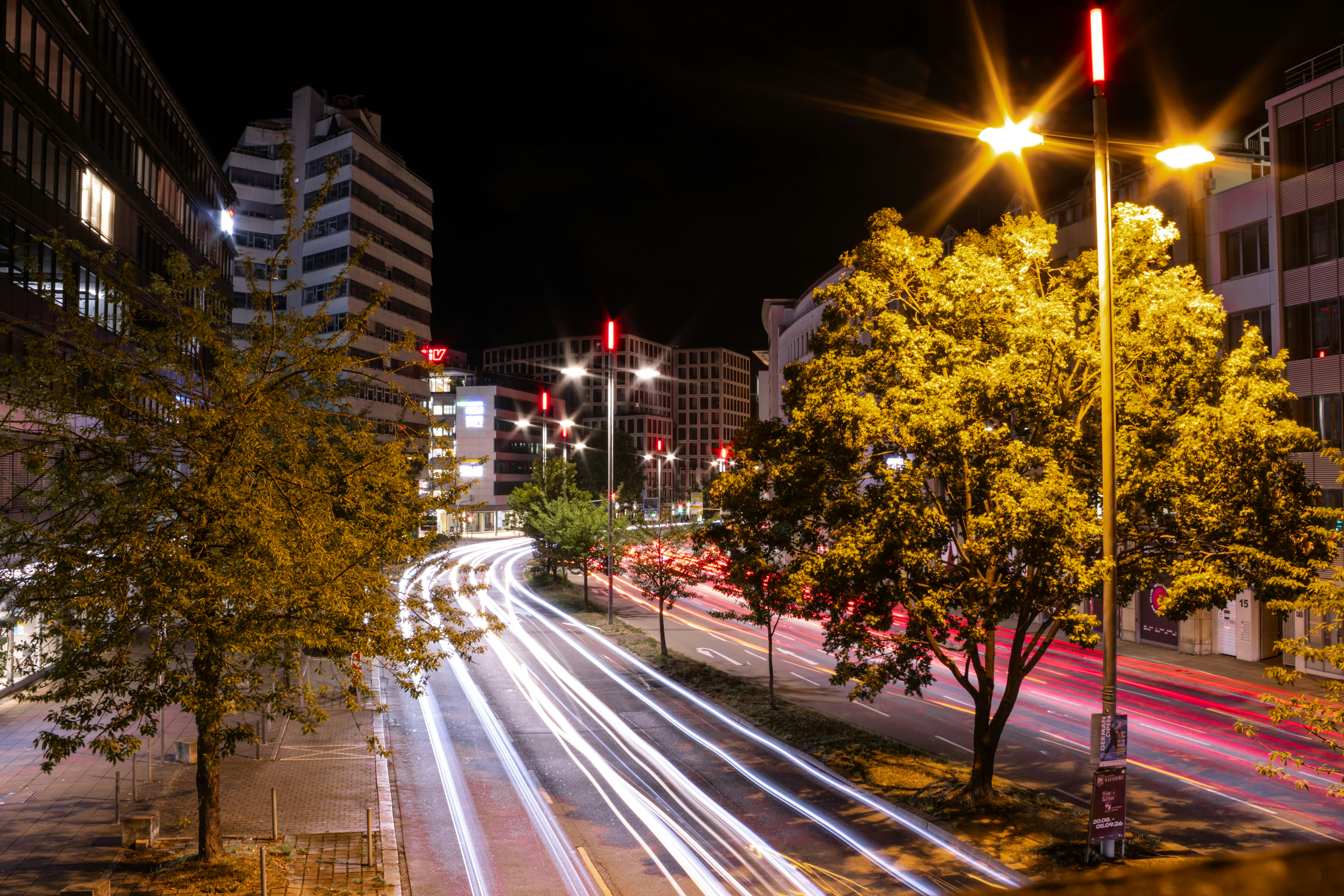 Long-exposure urban night scene with light trails from moving vehicles along a tree-lined street and modern buildings in the background.