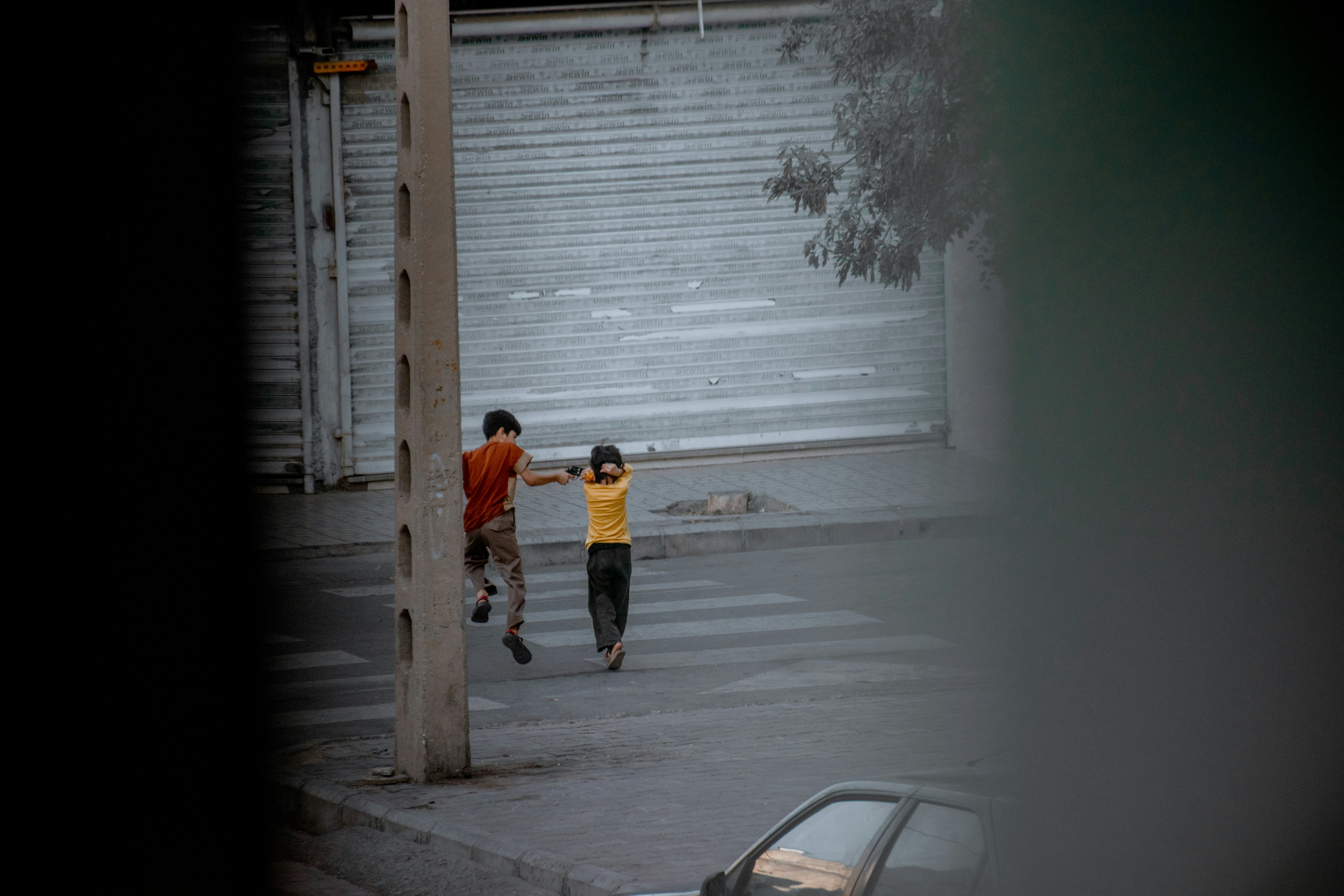 Two Children in a Quiet Street Reach for Each Other Behind a Parked Car