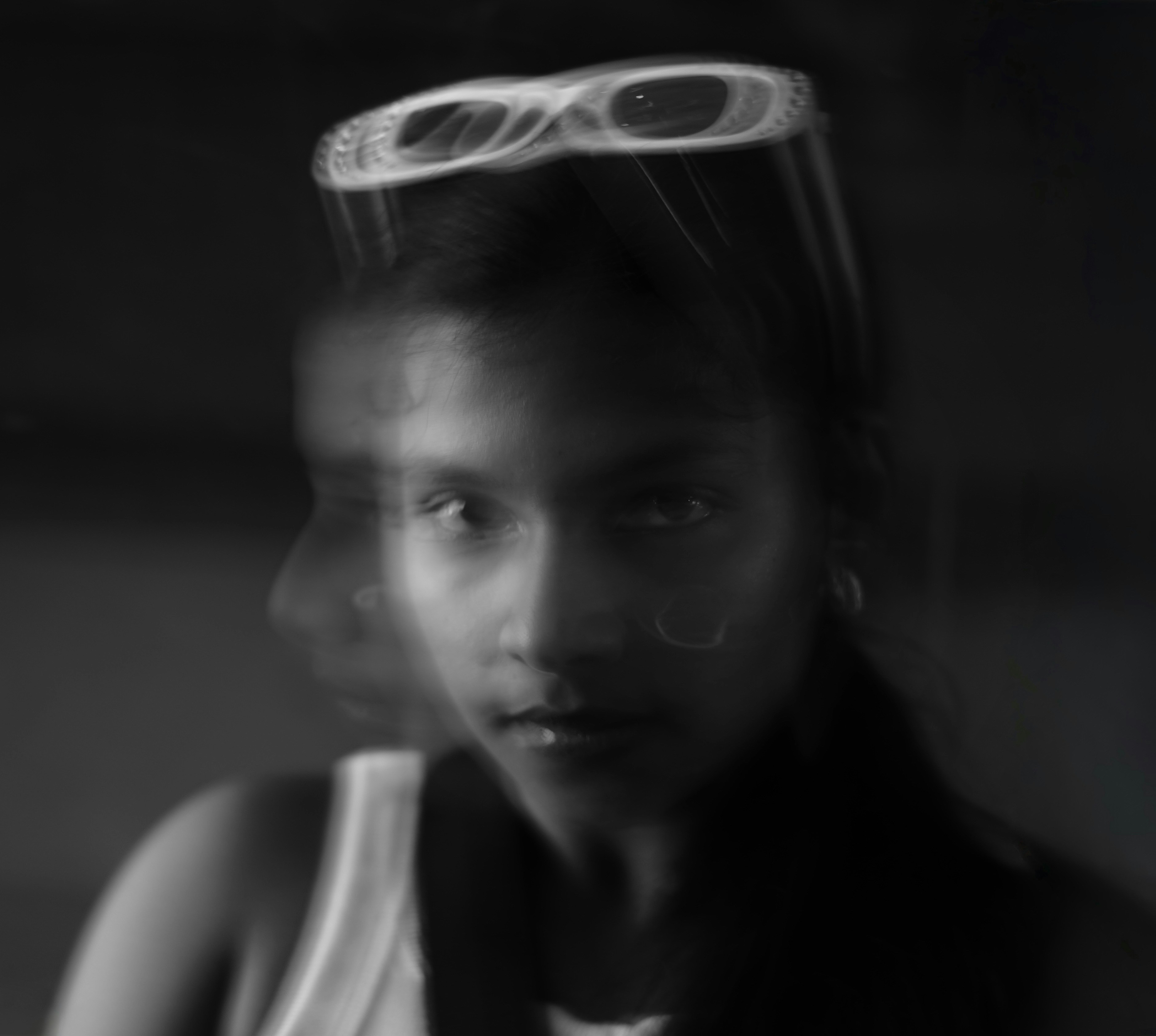 A softly blurred portrait of a girl with glasses resting on her head, captured in grayscale with movement creating ghosted edges. The composition centers the face while conveying motion blur and introspective mood.