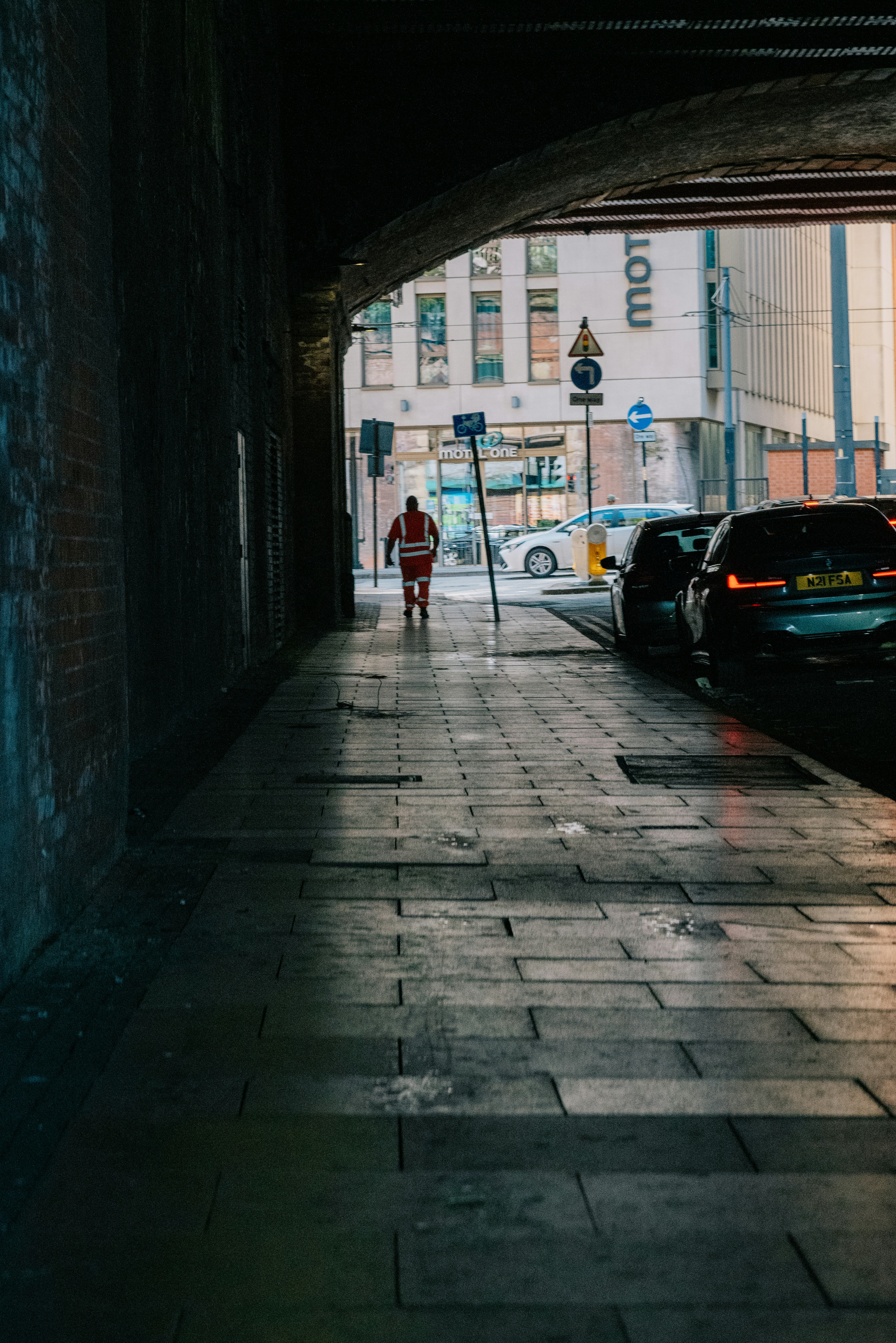 Site under a brick archway shows a wet, reflective sidewalk leading toward a distant figure in red, with cars and modern buildings beyond; the scene captures urban movement and atmosphere in a single frame.