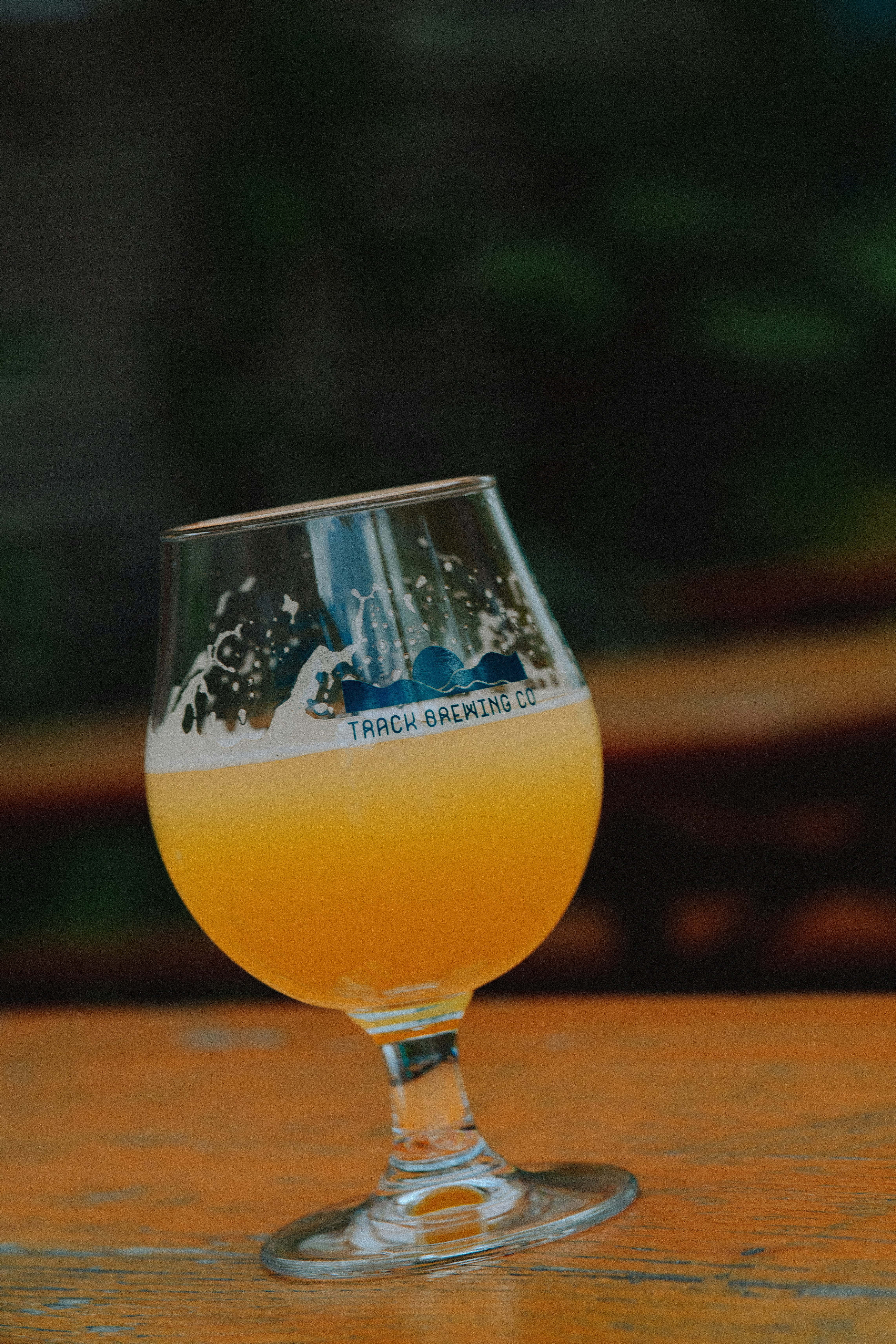 Glassy tulip glass with hazy orange beer sits on a wooden surface, foam remnants clinging to the rim; blurred outdoor background hints at a casual patio setting.
