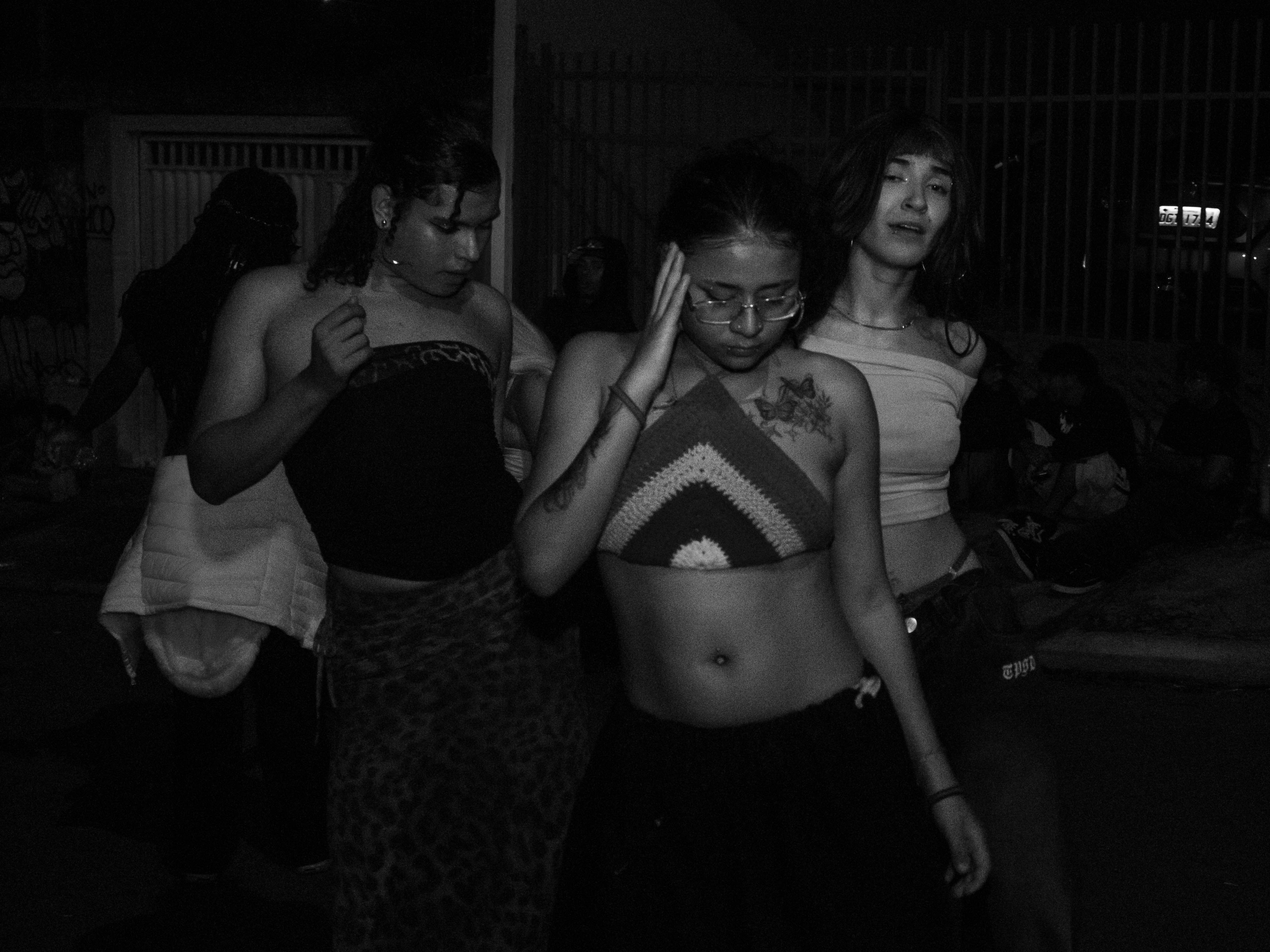 Three young women at night near a poolside pose candidly, one adjusting hair and another in a striped crop top; the grayscale scene emphasizes mood and group dynamics at a casual outdoor gathering.
