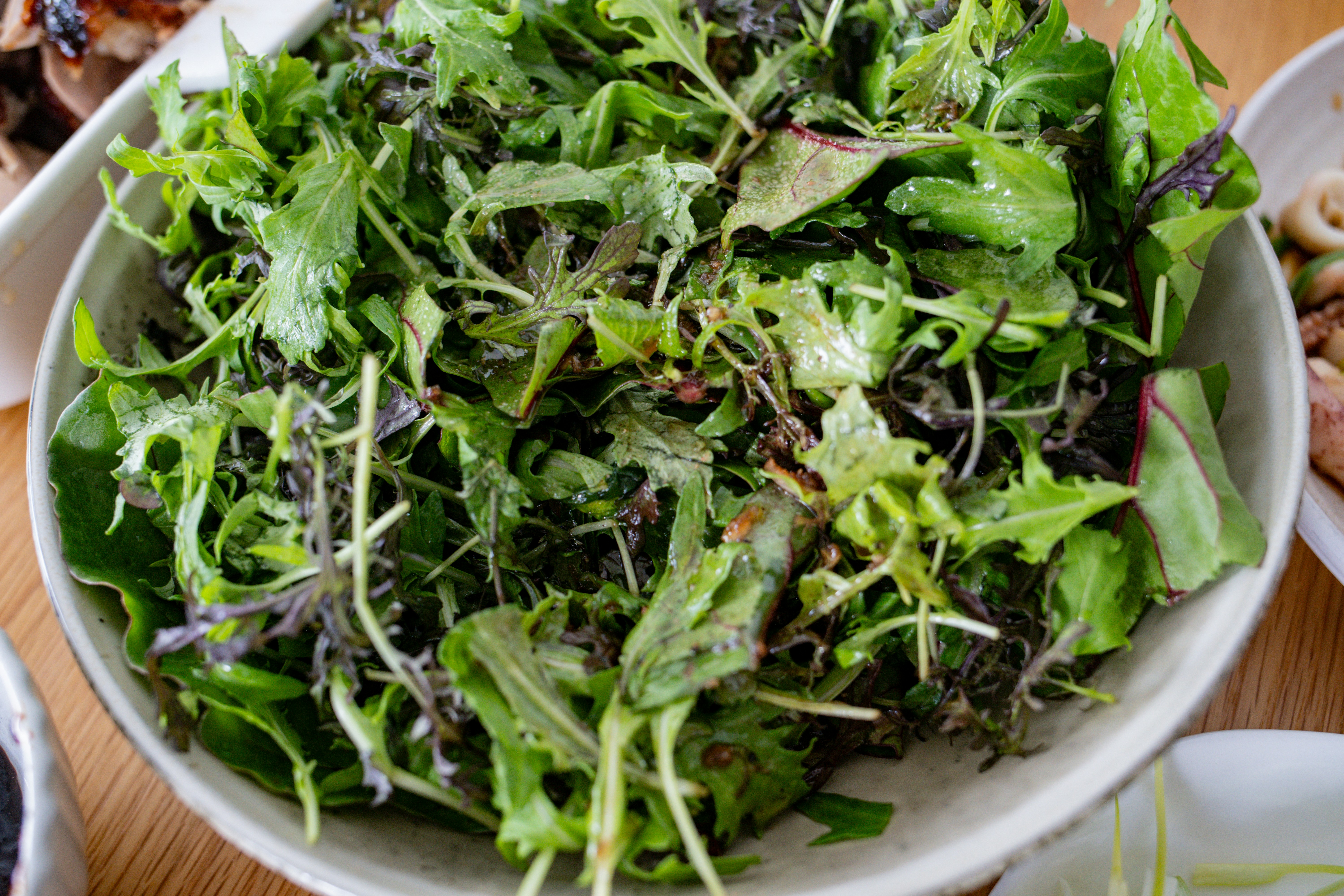 A bowl of assorted fresh greens laid atop a wooden table, showcasing vibrant leaves and stems. A healthy herb-and-veggie centerpiece meant for a meal or salad preparation.