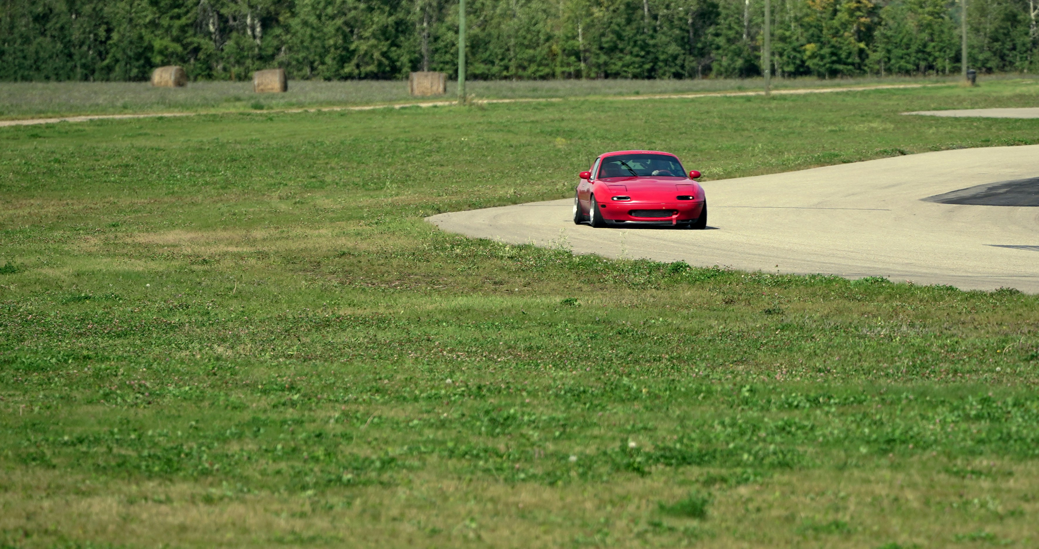 A bright red car negotiates a gentle curve on a light-tan road surrounded by expansive green fields.