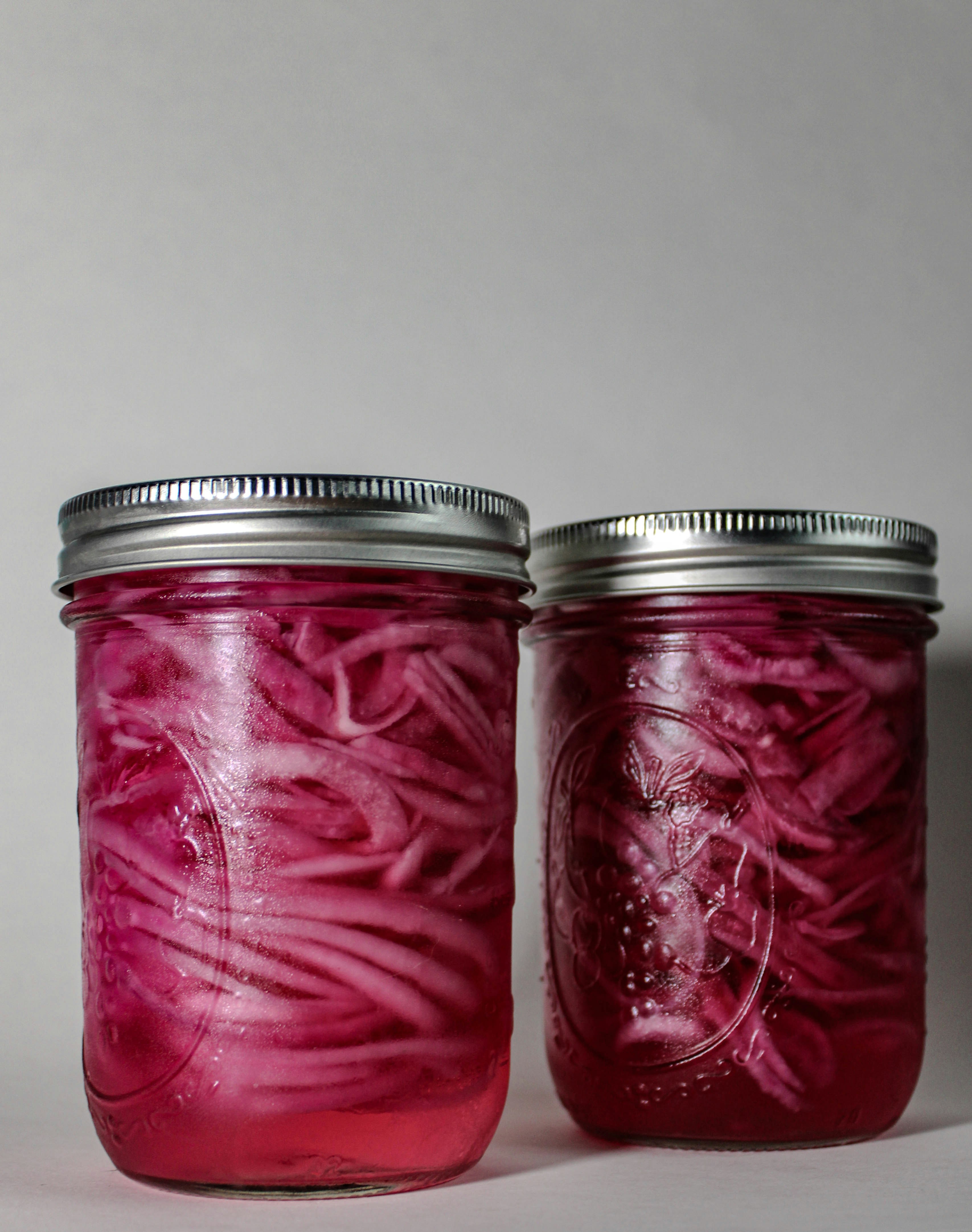 Two glass mason jars filled with bright pink pickled noodles sit on a neutral surface, the left jar slightly larger and lighter, creating a subtle contrast in texture and color.