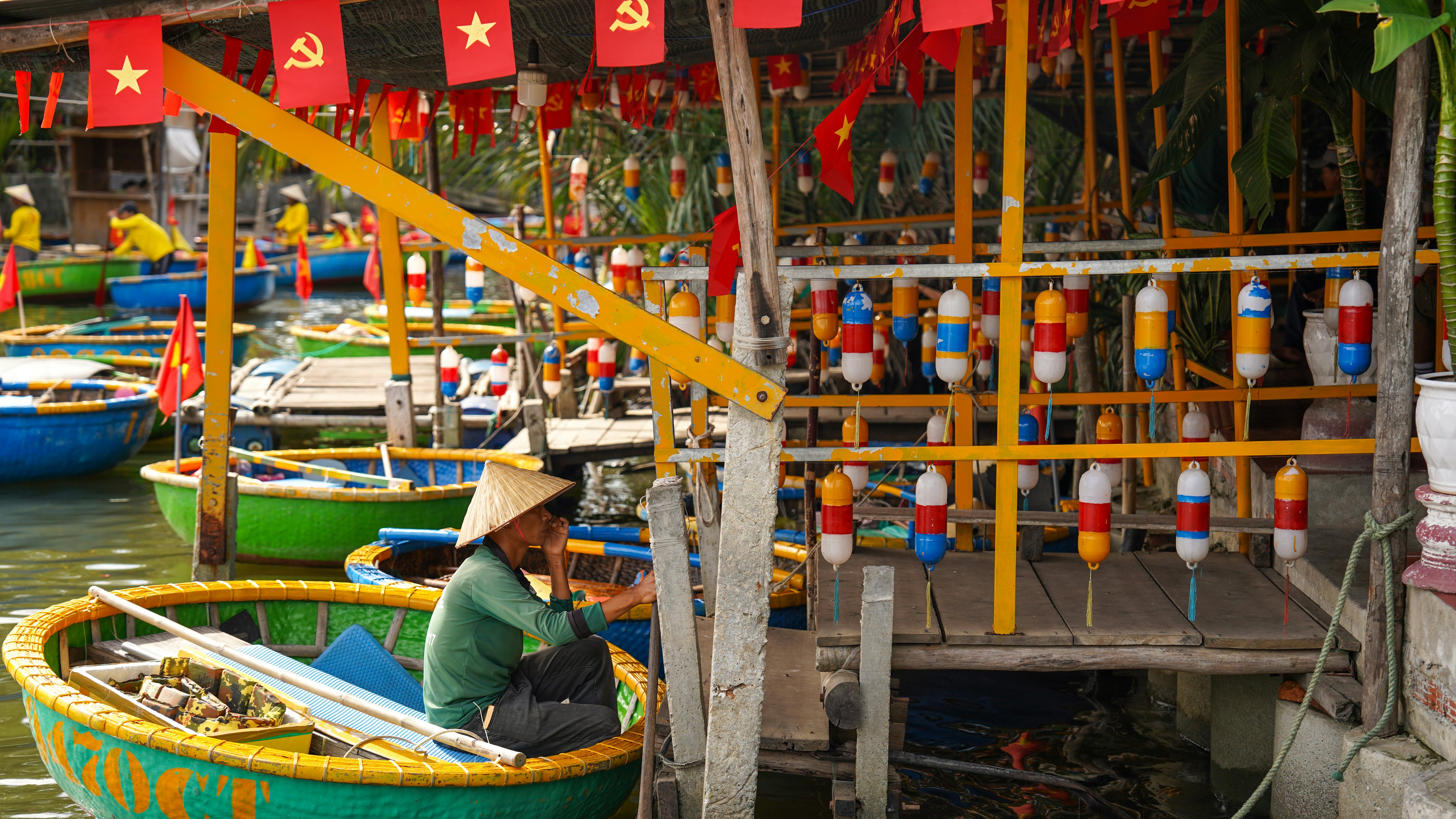 Colorful boats and a wooden dock with red flags and buoys; a fisherman sits in a small boat by calm water at a riverside market.