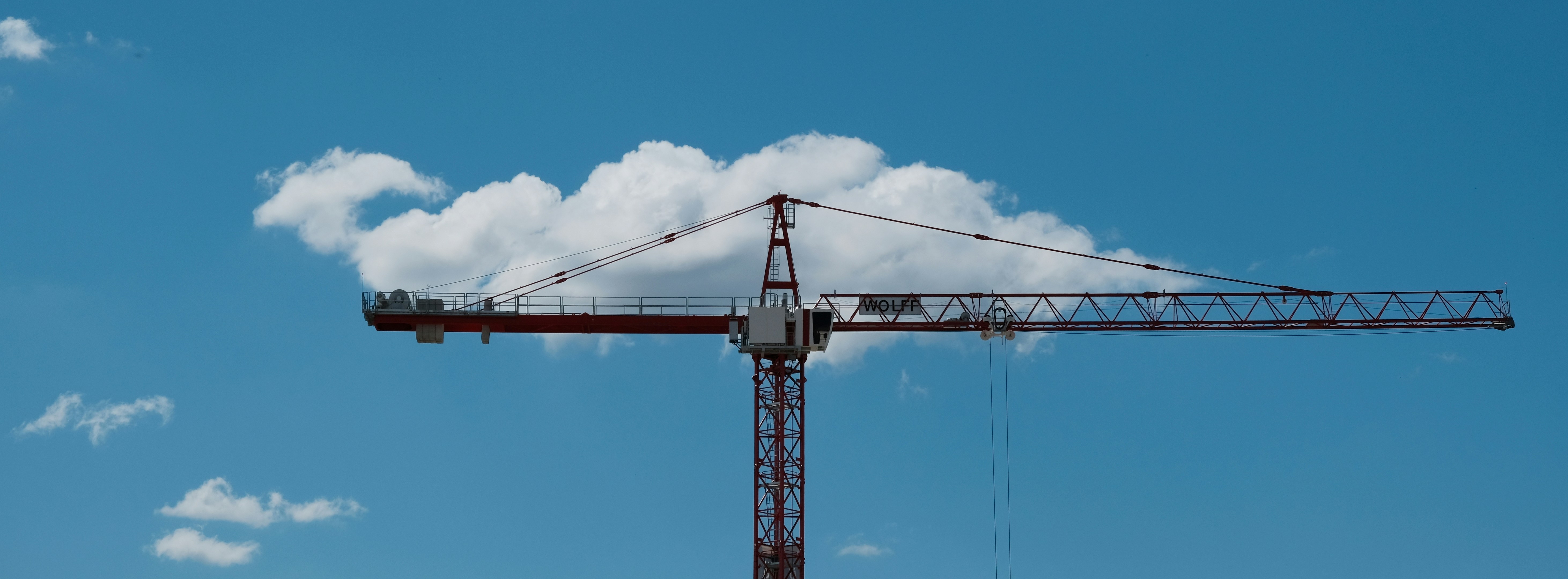 Tall construction crane spans a bright blue sky with wispy clouds, its red framework forming a strong horizontal line and a quiet industrial focal point.