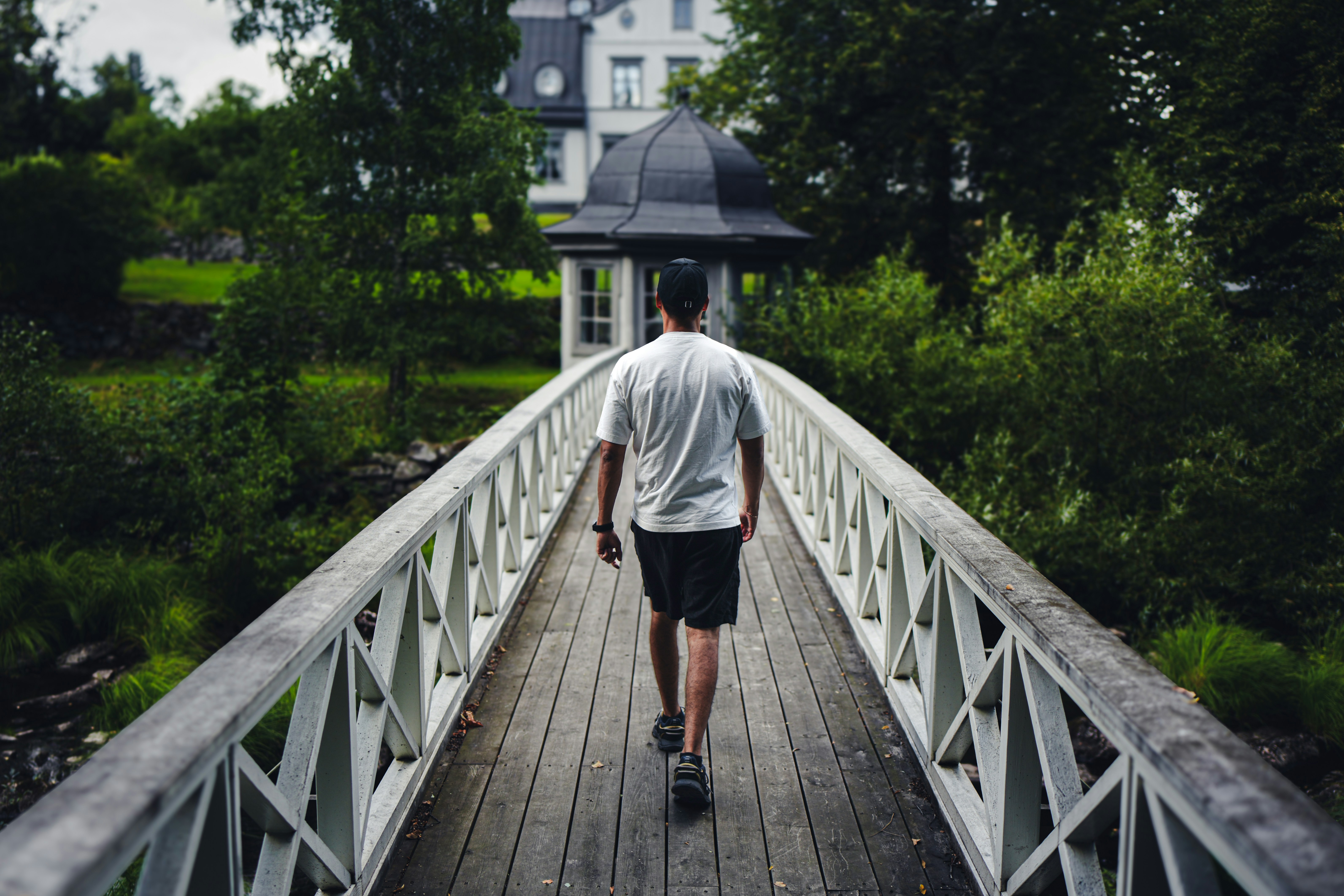 A man walks away across a white-panel wooden bridge toward a quaint pavilion, surrounded by lush greenery and a tranquil landscape.