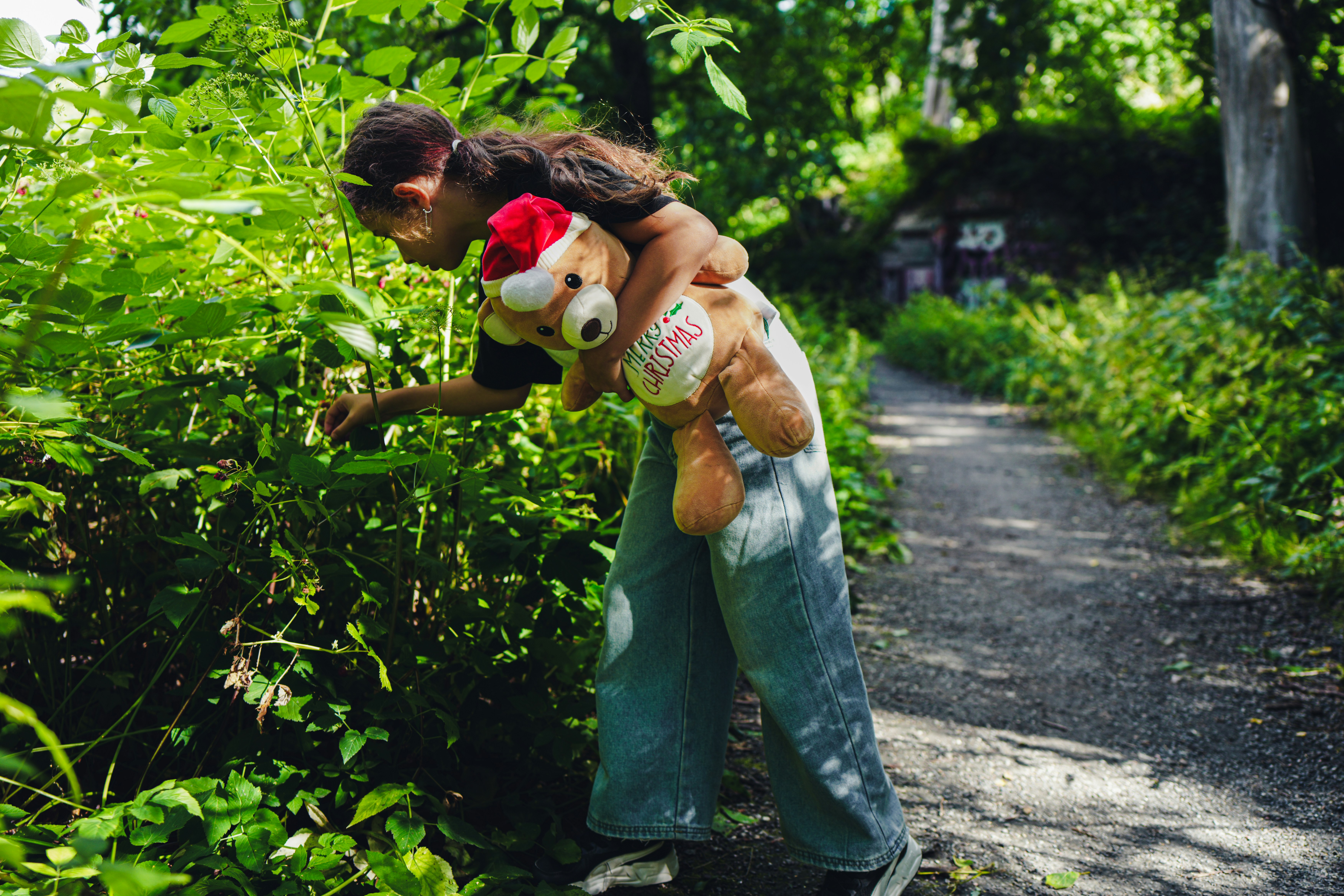 Child with stuffed bear bends to examine foliage on a sunlit garden path