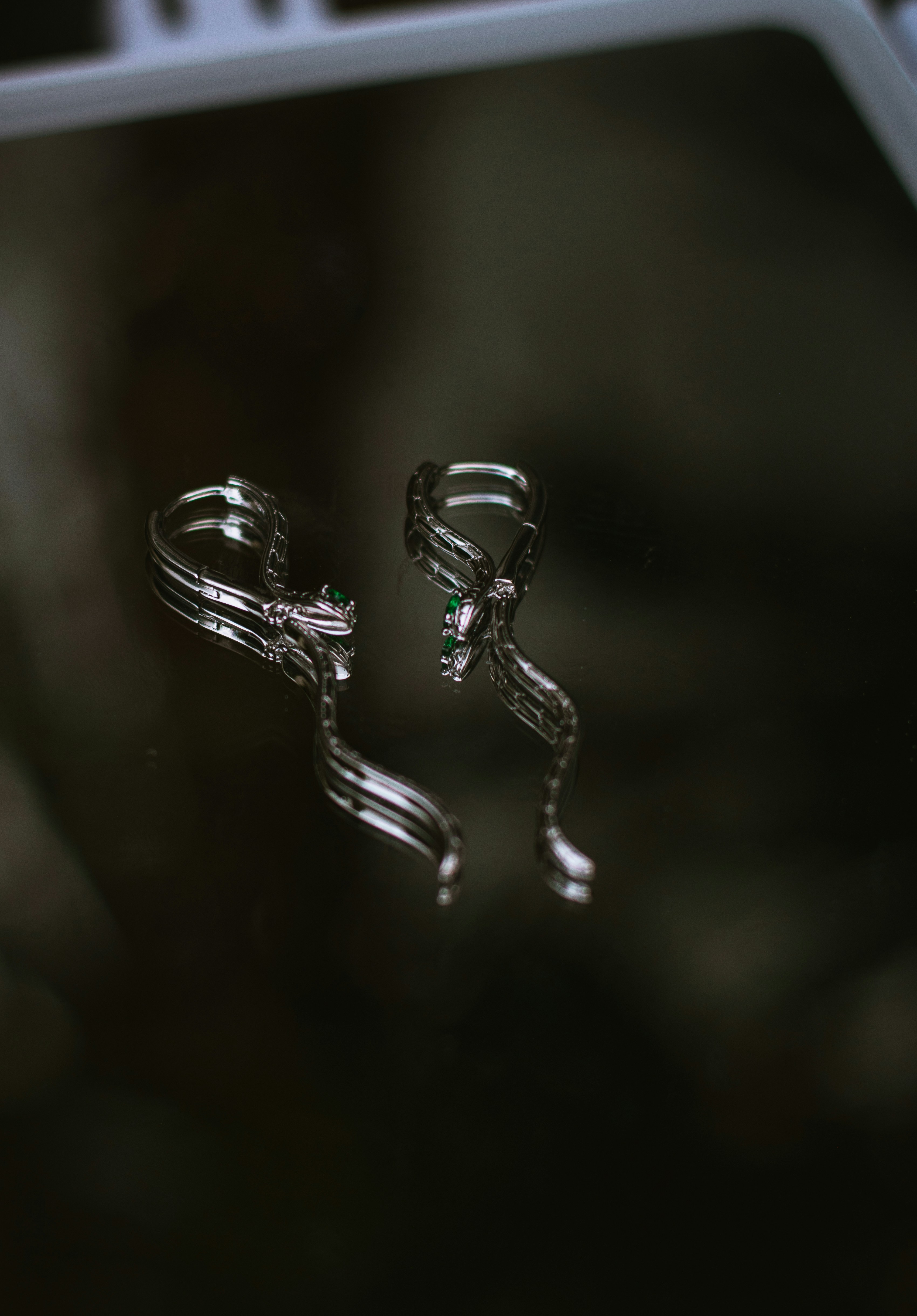 Gleaming Double Spirals: Emerald-Accented Rings Resting on a Reflective Surface