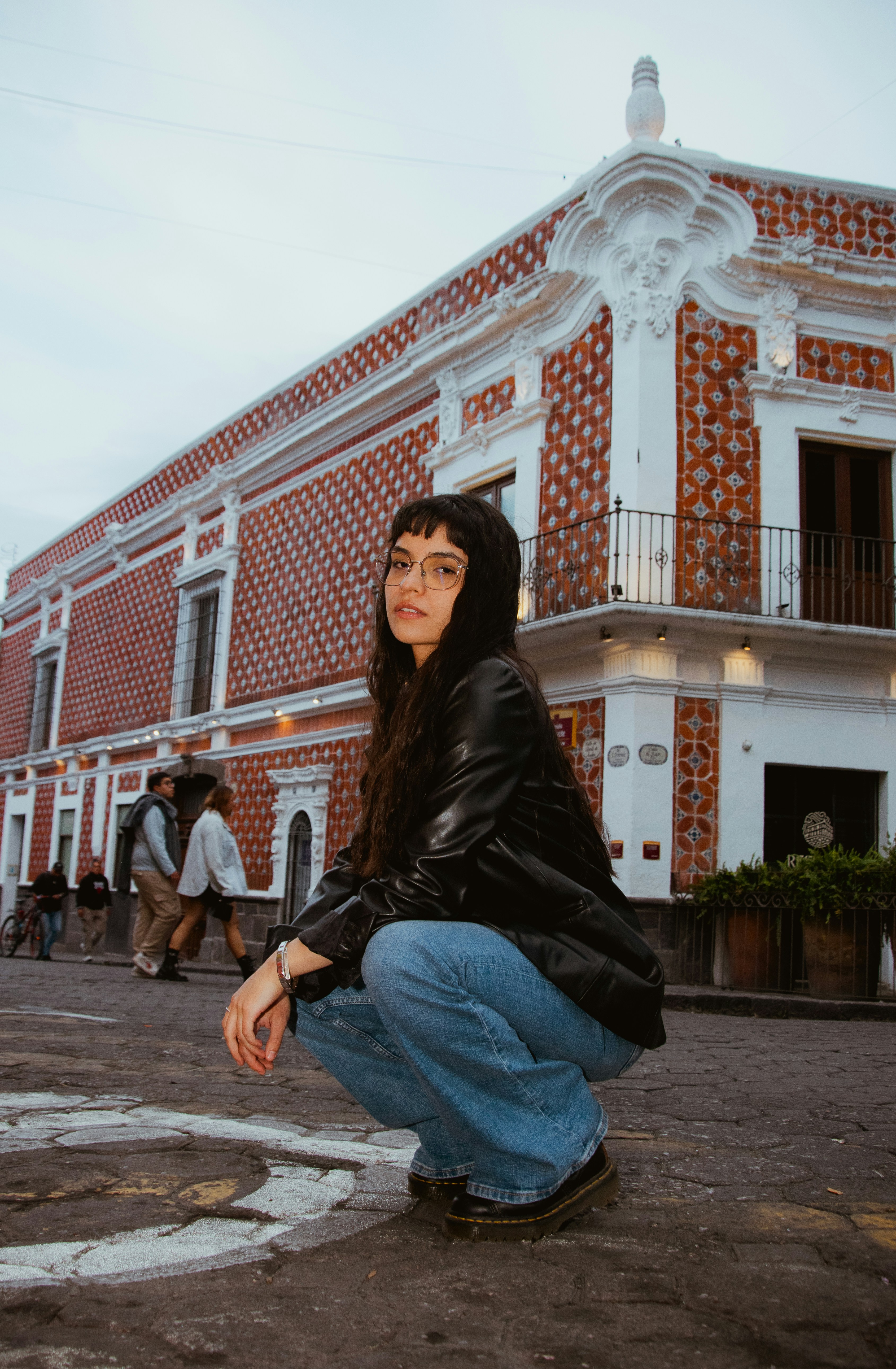 Woman in leather jacket crouches before a patterned colonial building