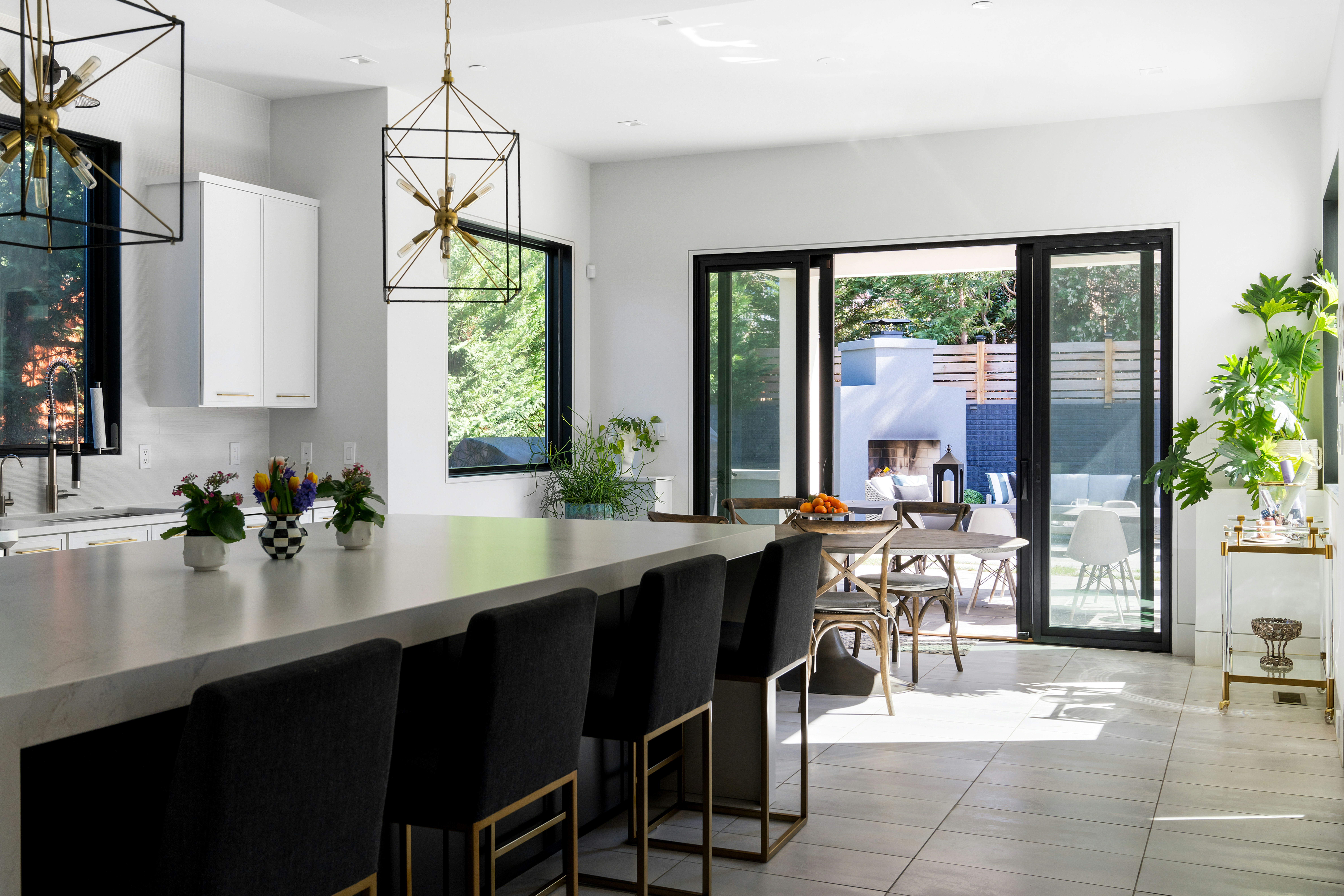 Bright open kitchen with a long island and bar seating, leading to a glass-paned dining area and outdoor space; clean lines, greenery, and ample natural light create an airy, contemporary home setting.