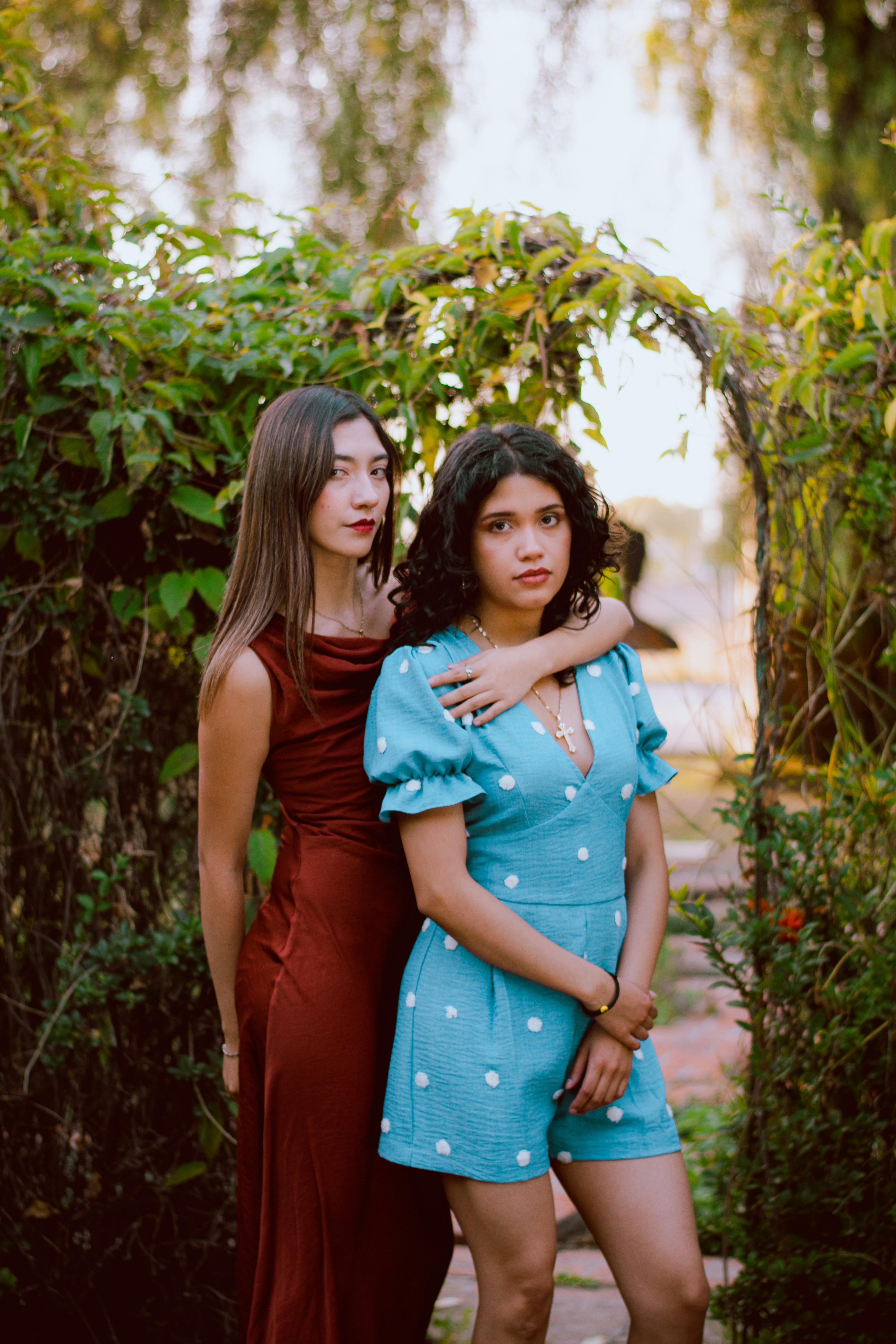 Two friends in a sunlit archway, contrasting dresses and a soft, leafy backdrop
