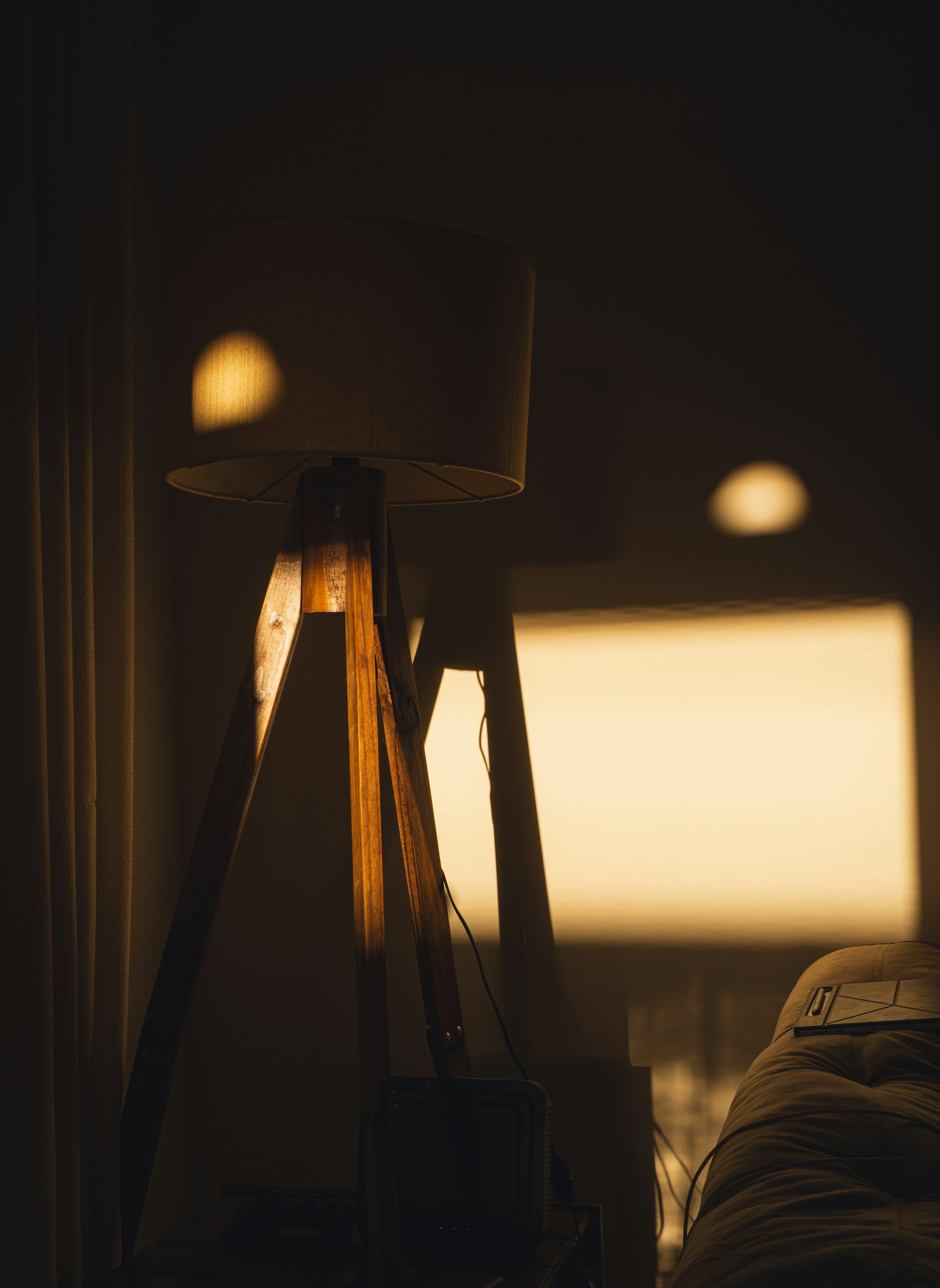 A wooden tripod floor lamp stands in the warm glow of evening light, framed by curtains and the soft geometry of a lit window. A quiet room at the end of the day.