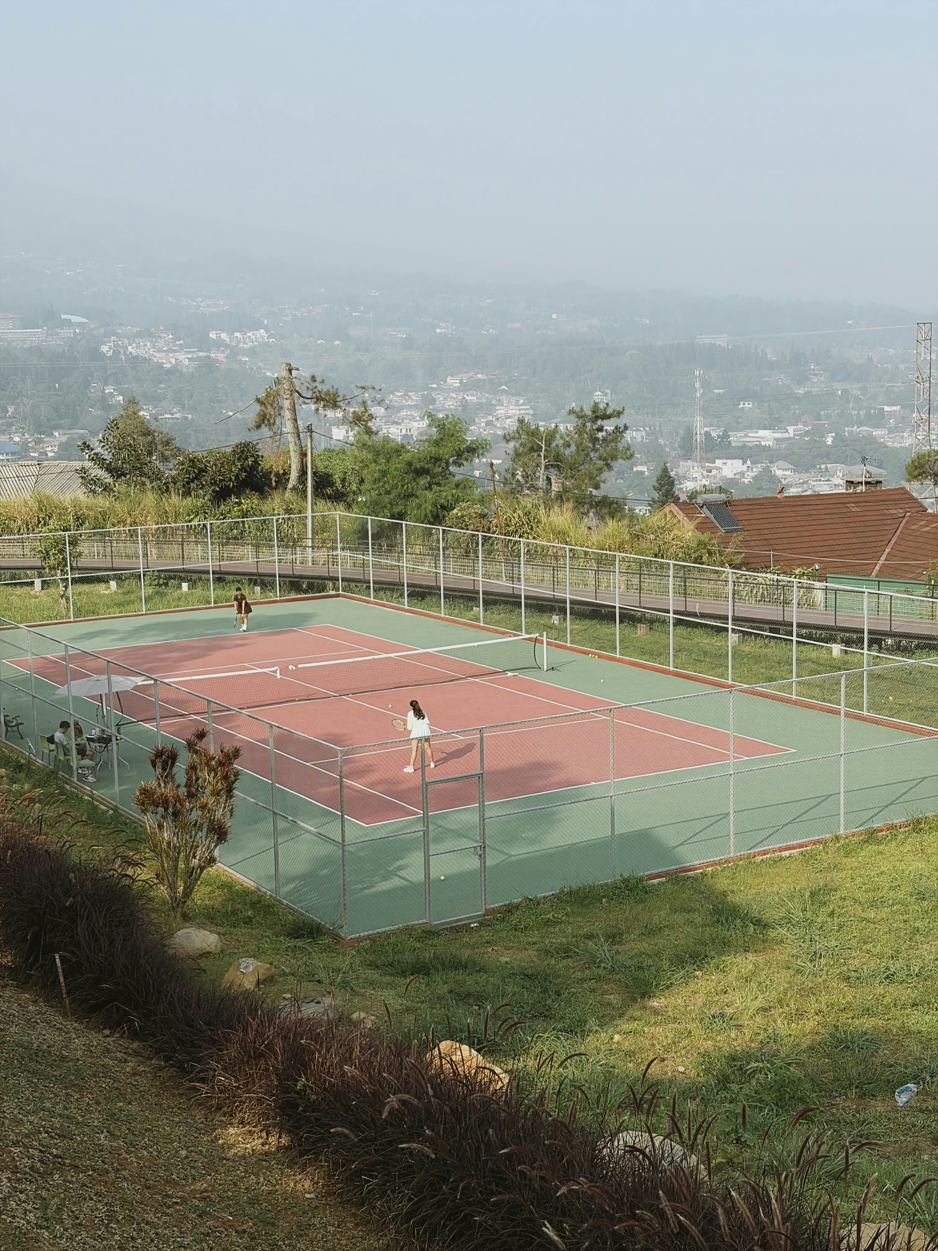 An elevated tennis court tucked in a hillside with players on court and a distant cityscape blurred by mist.
