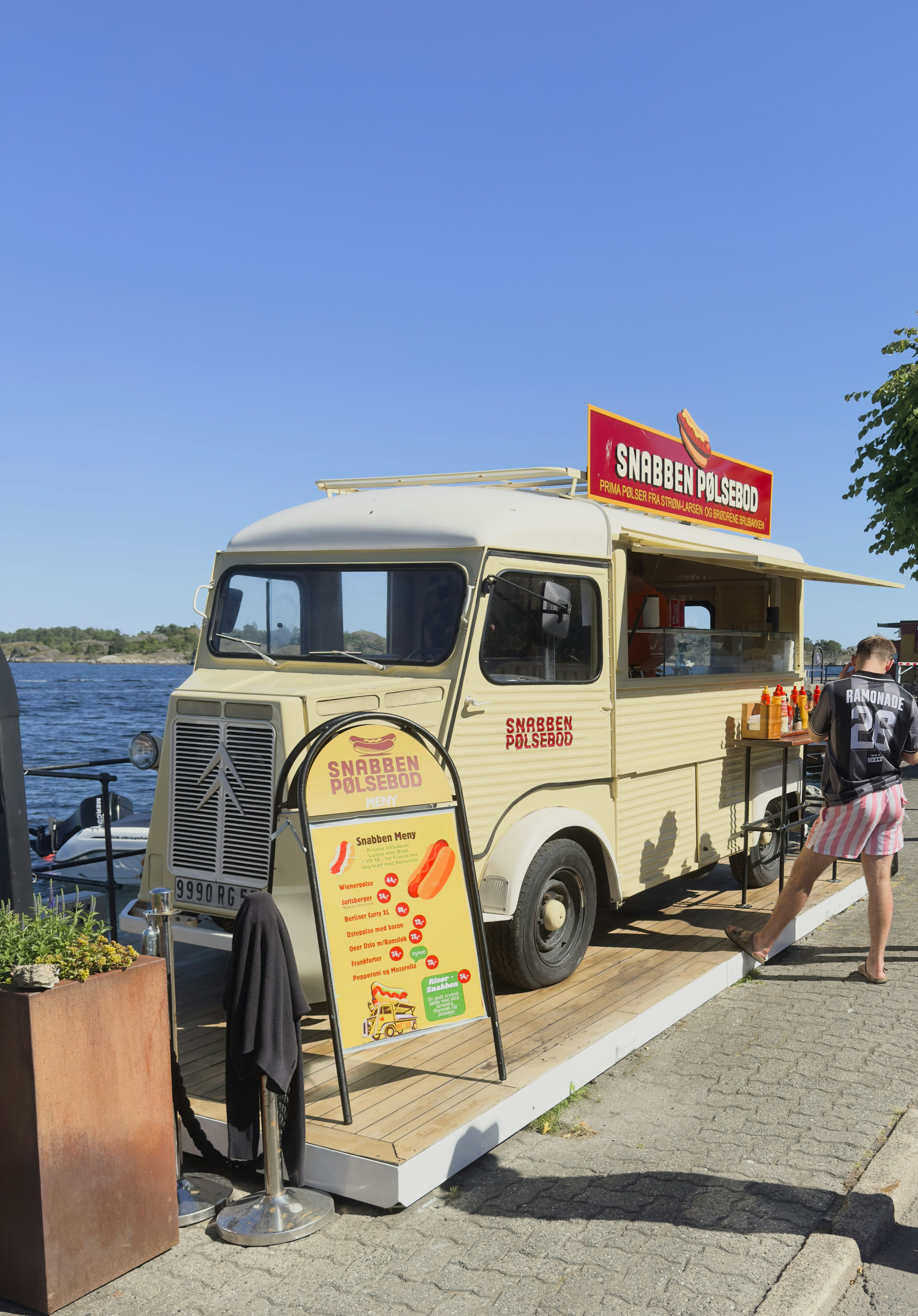 A retro beige food truck serves customers by the water with a menu board and a person at the side; a bright, sunny day highlights the scene.