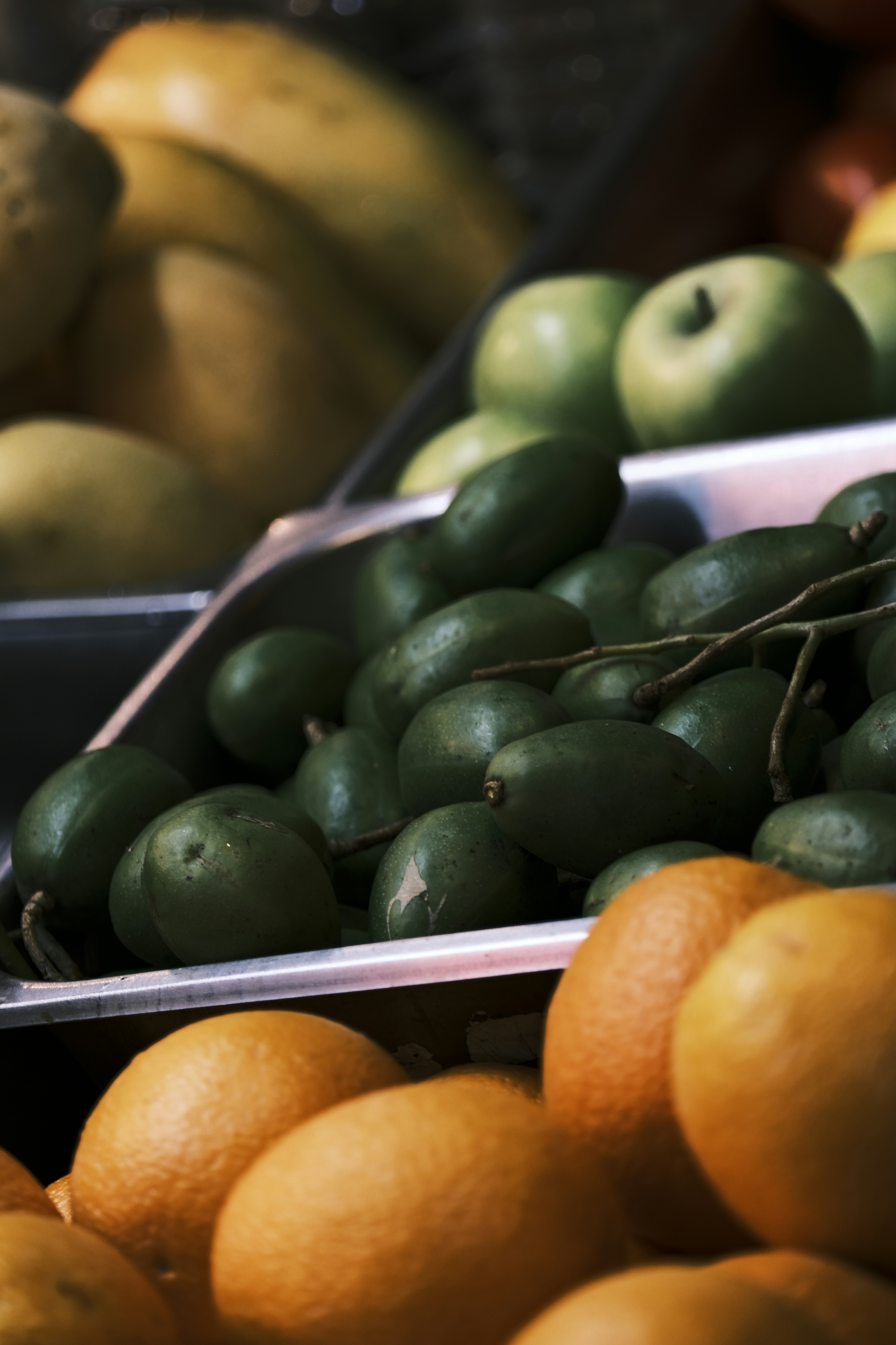 A metal tray of green hog plums beside oranges and green apples.