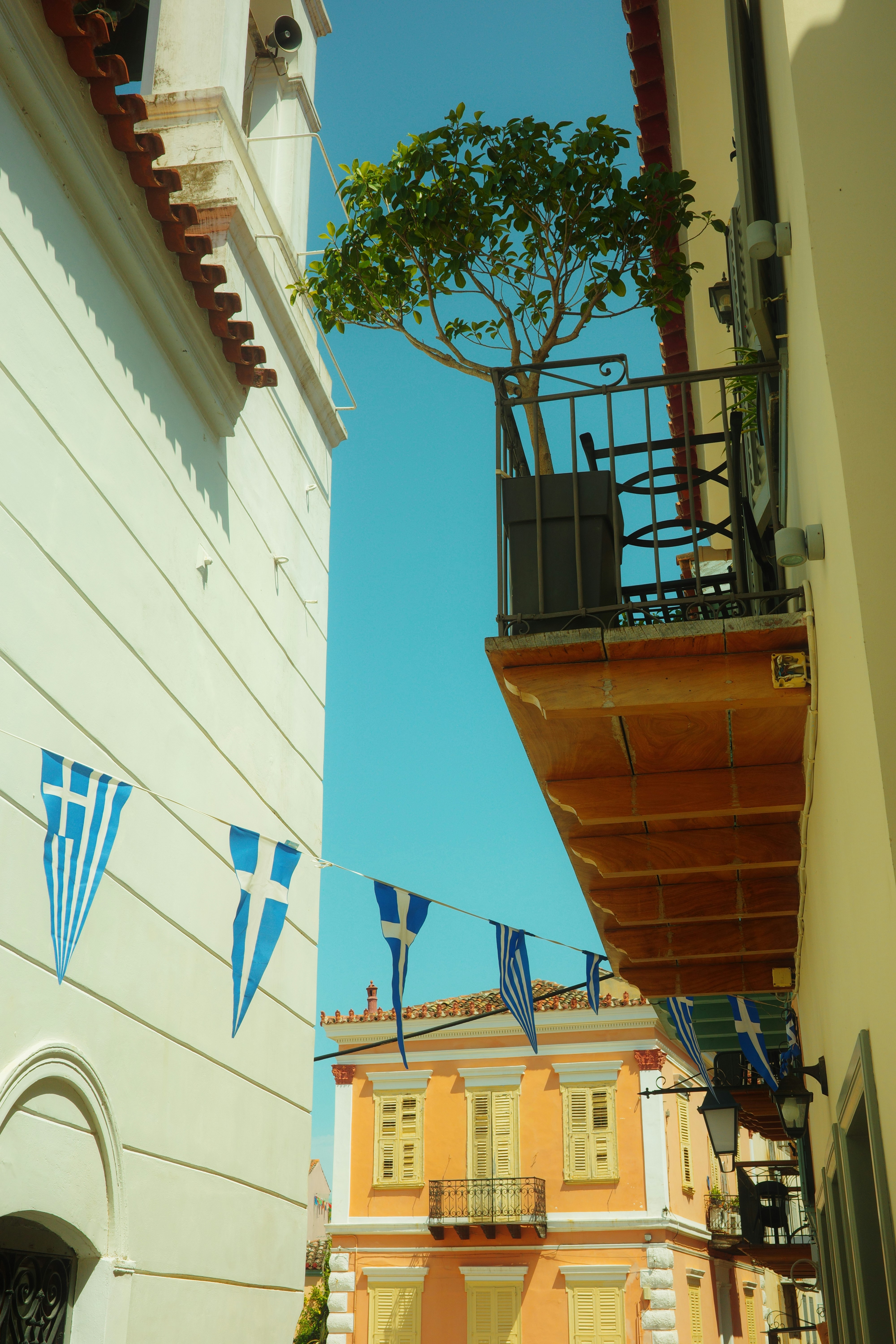 Greek flags hanging between buildings on a narrow street under a clear blue sky.