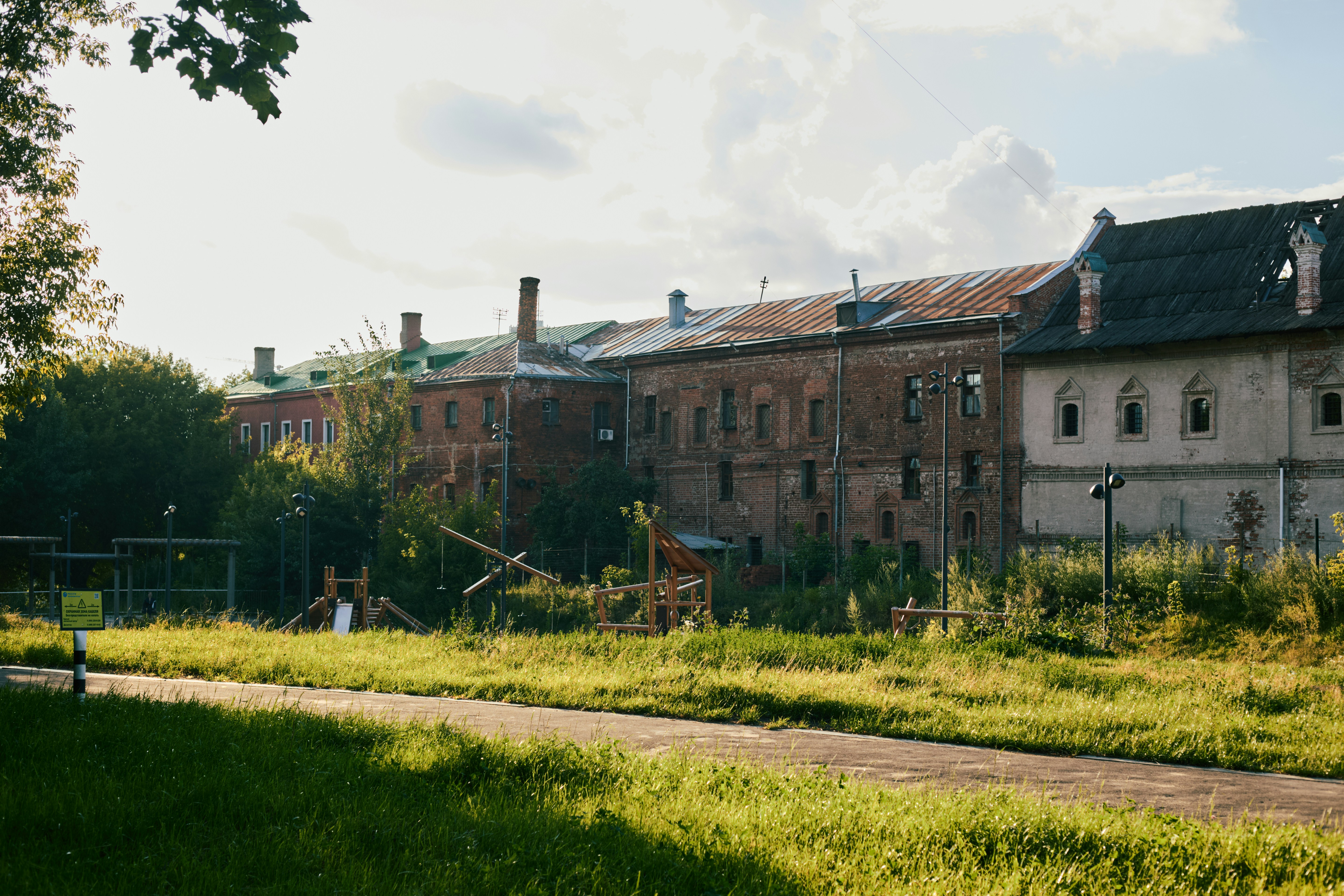 Sunlit brick factory facade overgrown with grass and distant playground structures